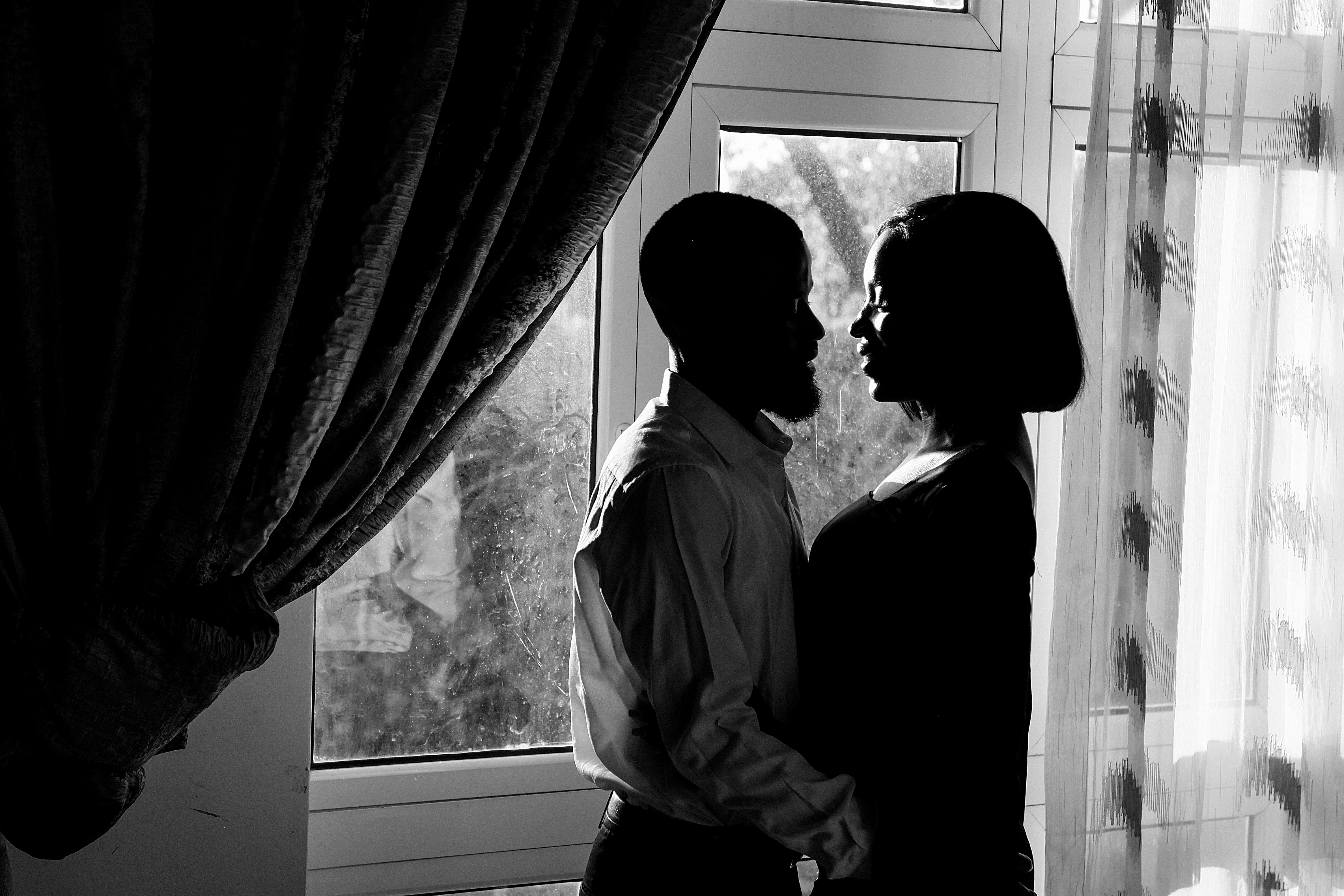 A couple embraces in silhouette against a softly lit window, framed by elegant curtains. The scene captures an intimate moment of connection.