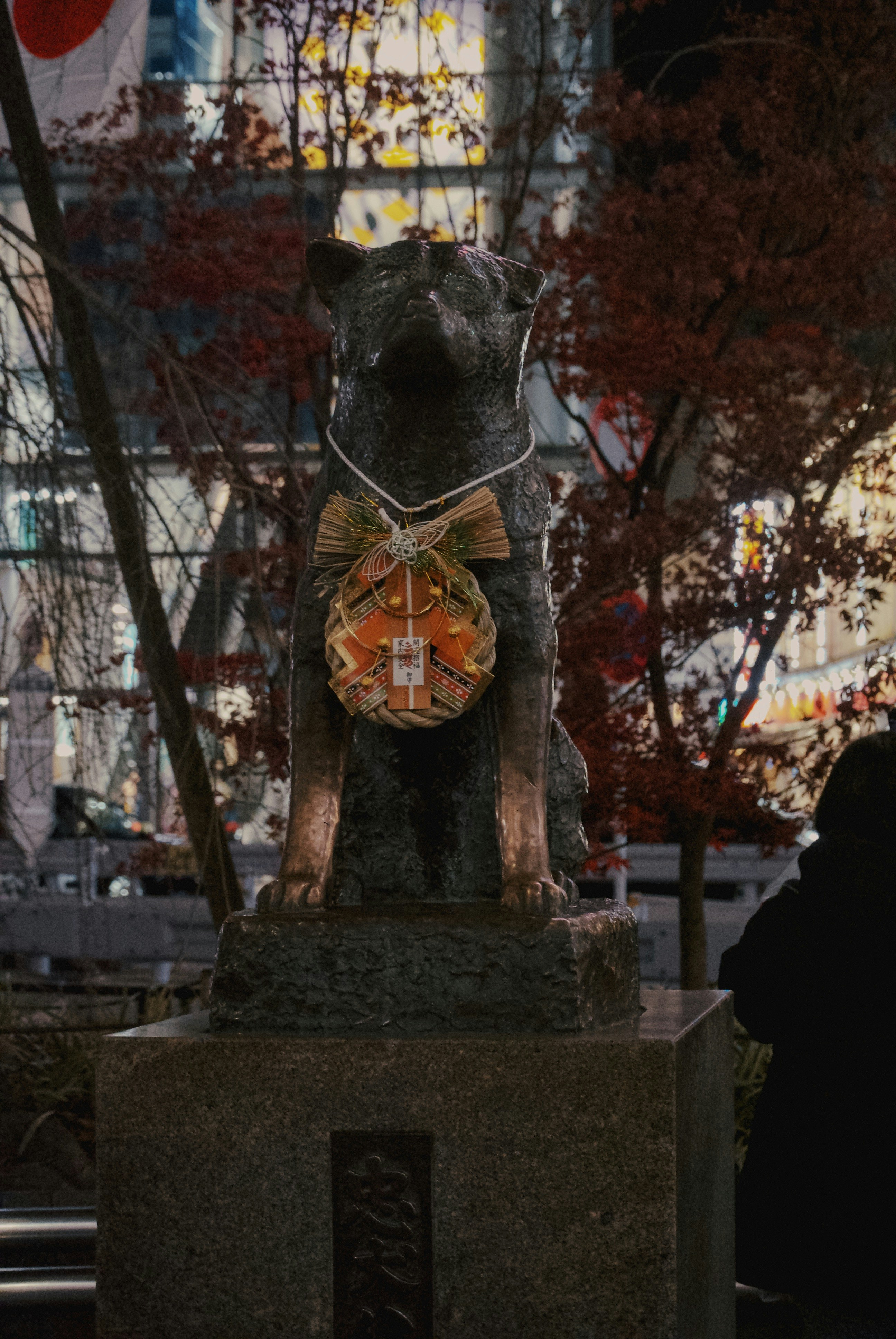 Stone guardian dog statue at dusk, adorned with a ceremonial bib and sacred amulets; red maple leaves and city lights provide a moody urban backdrop.
