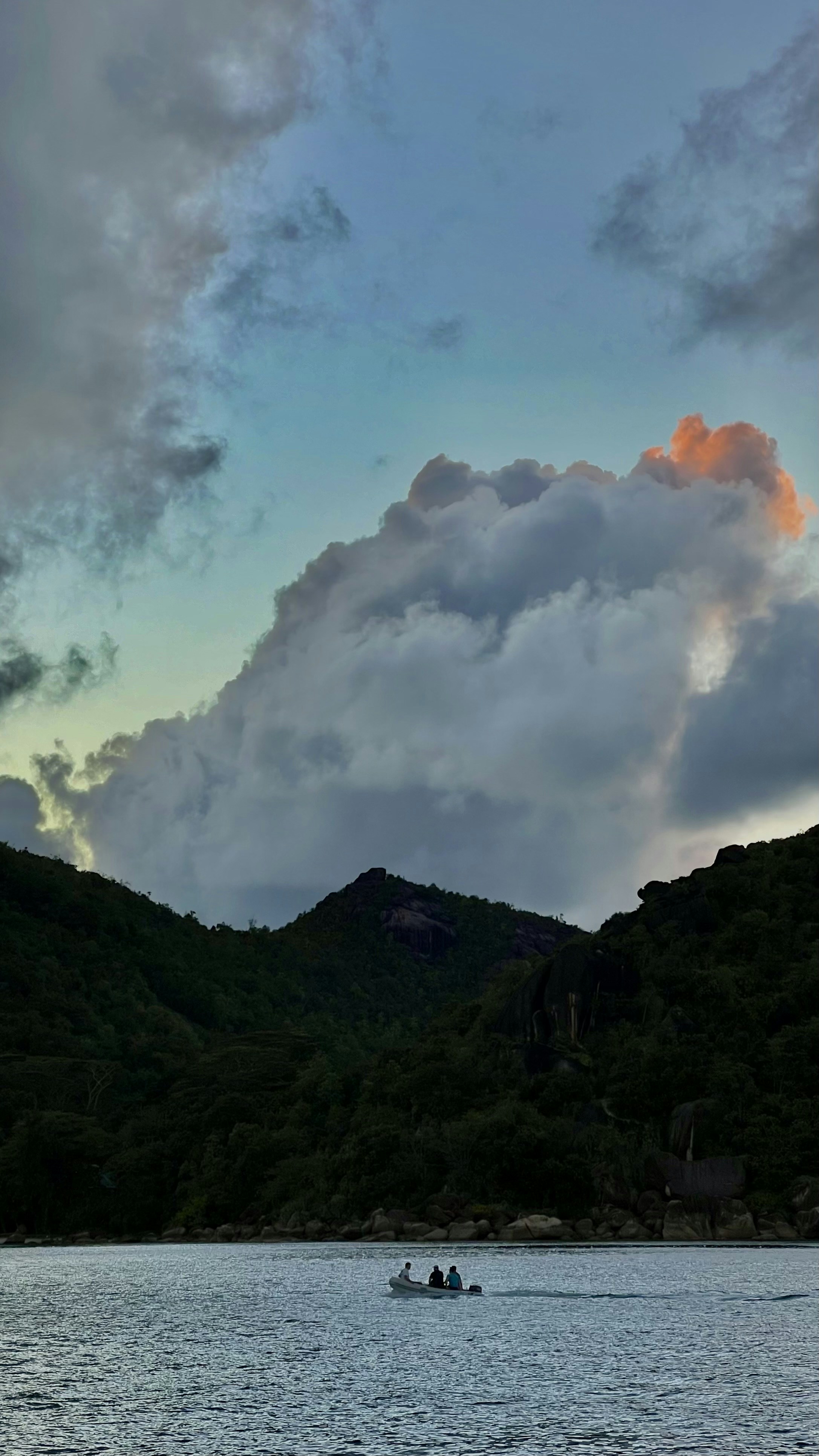 Small Boat Under Dramatic Twilight Clouds - A small boat glides over calm waters, surrounded by lush hills and dramatic clouds tinged with sunset colors. The peaceful yet moody ambiance captures the transition from day to night. This image evokes a sense of solitude and connection with nature.