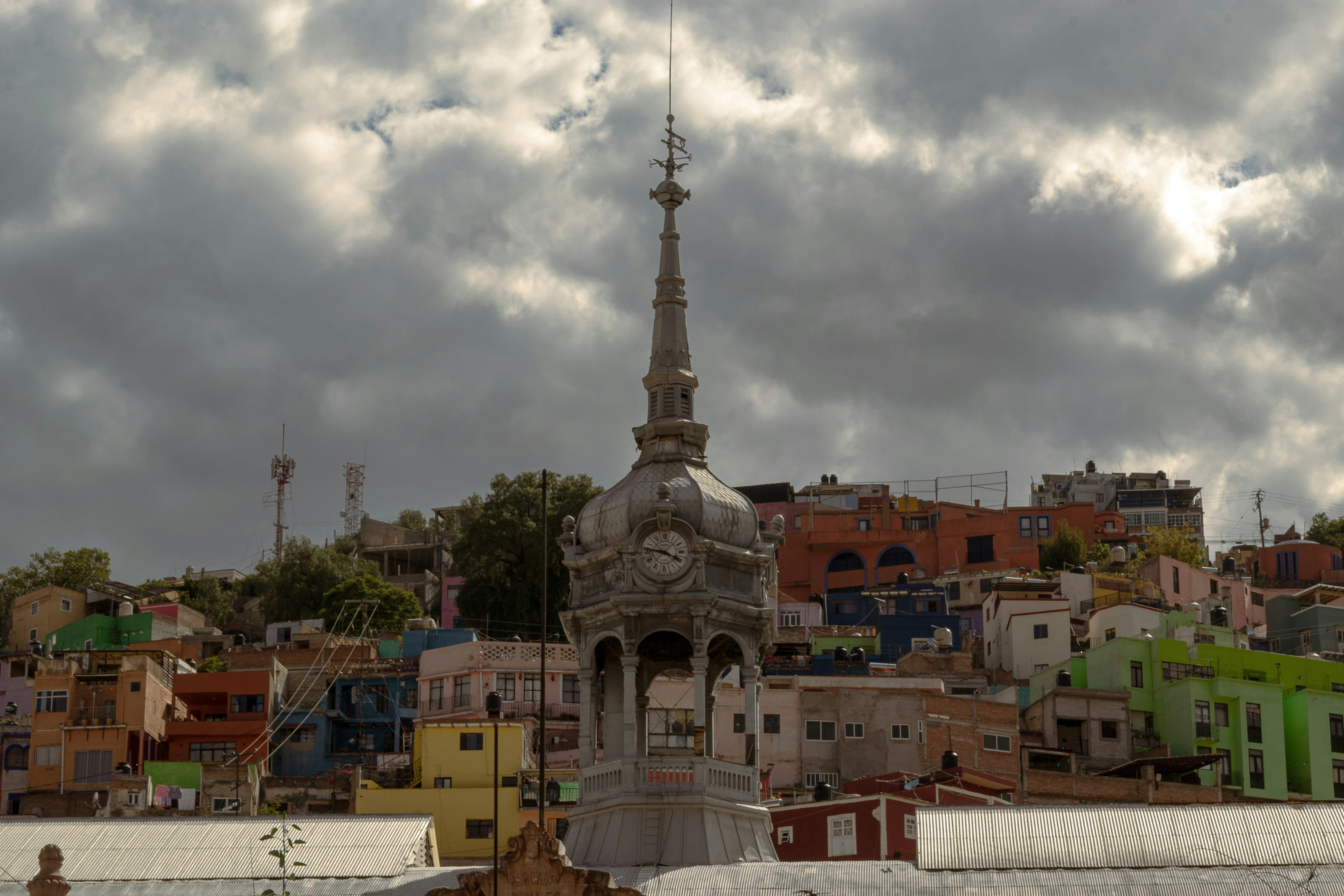 Clock-topped dome rises before colorful hillside homes under a moody, cloud-filled sky.