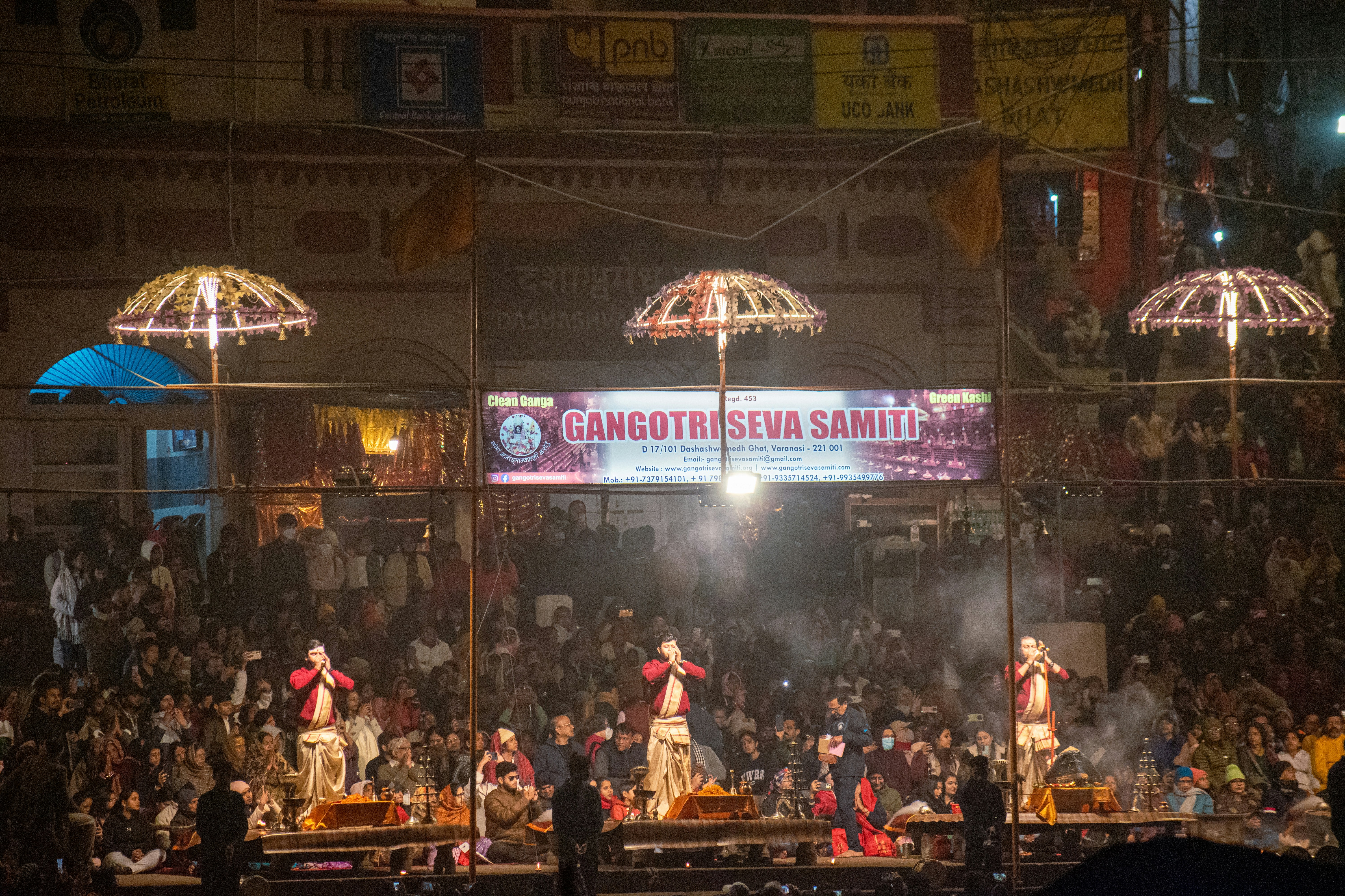Ceremonial performers on a lit stage surrounded by a large crowd at night.
