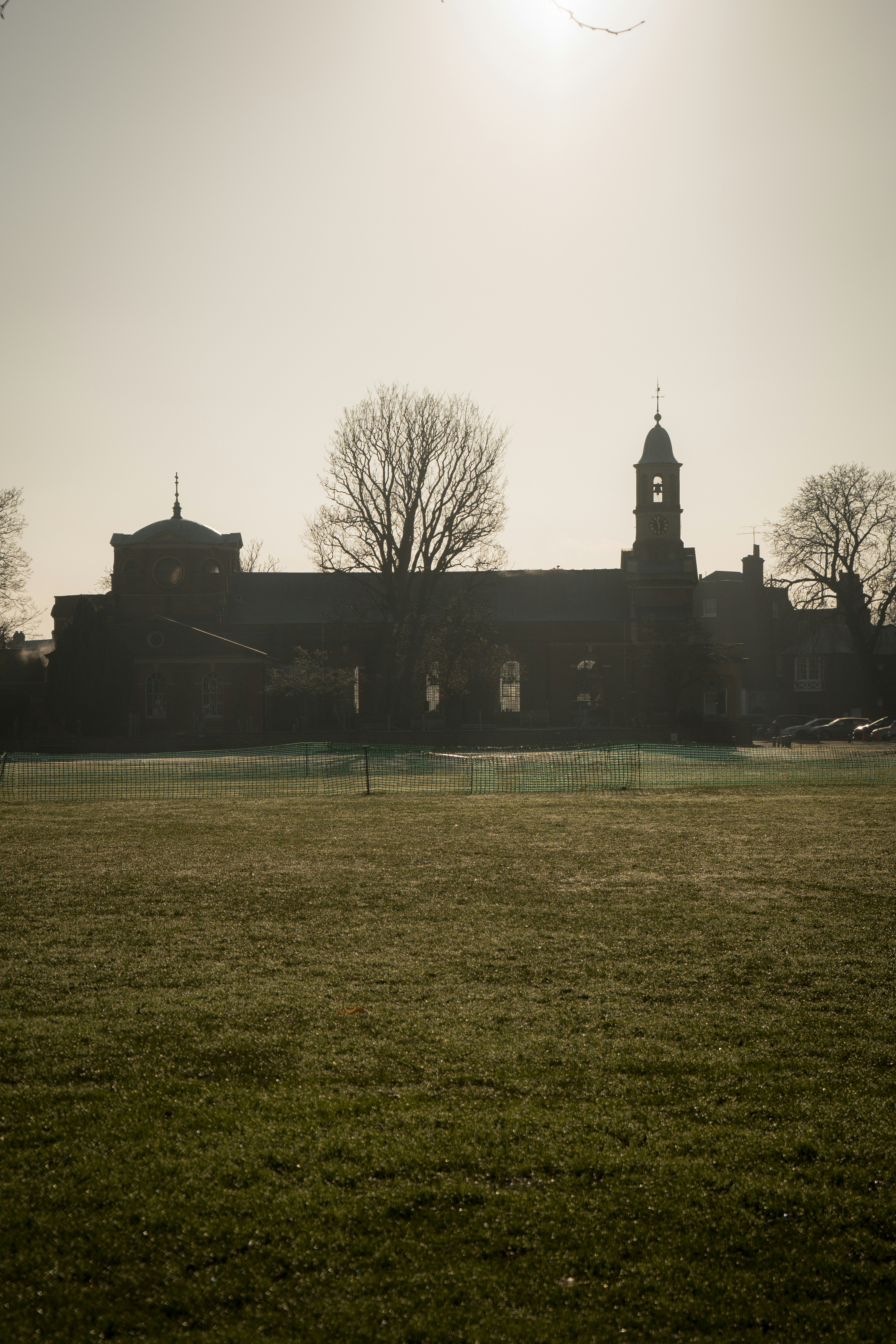 Silhouetted historic building framed by bare trees, set against a hazy morning sky. The tranquil park scene invites reflection.