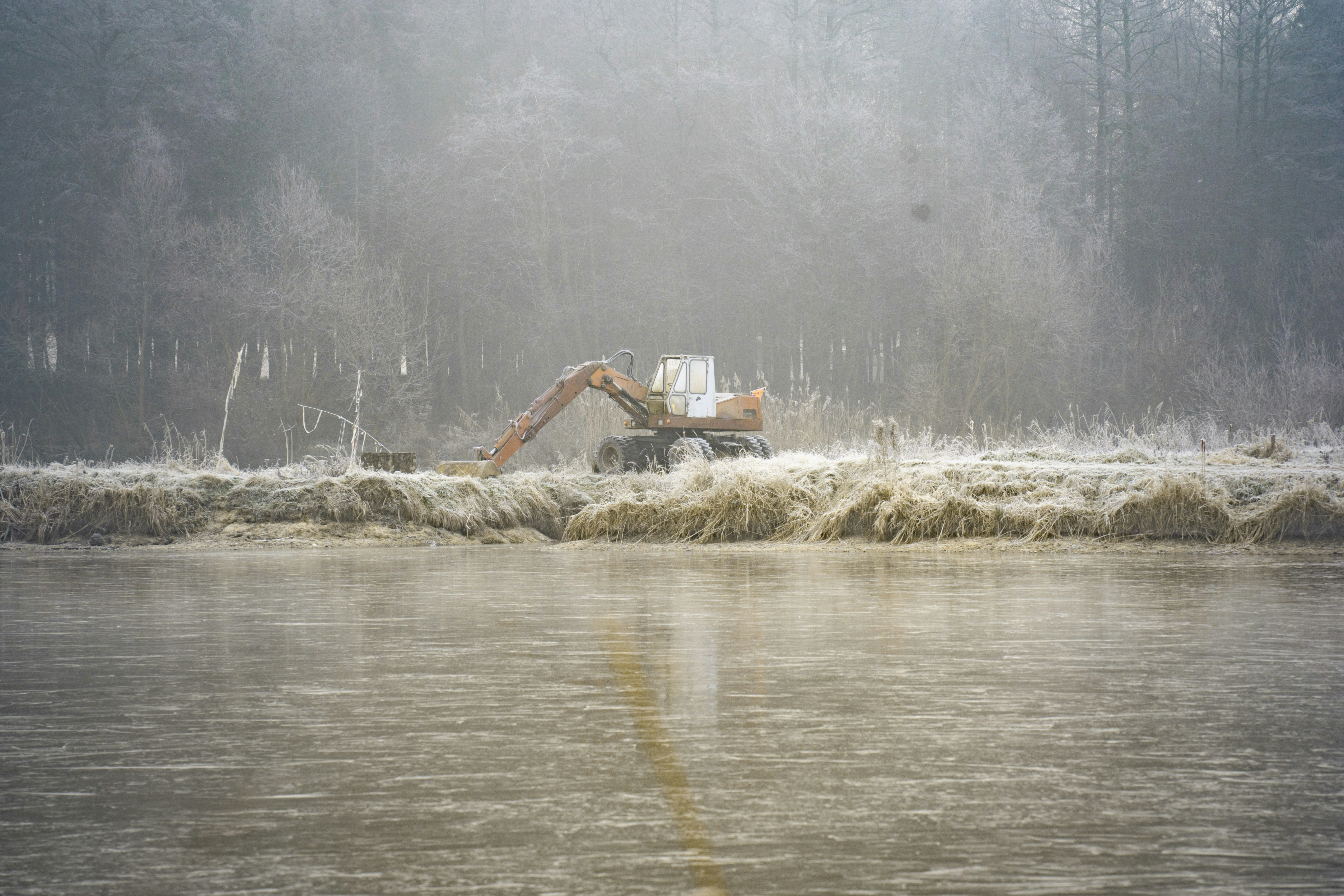 Excavator rests beside a tranquil frozen lake surrounded by frosty trees.