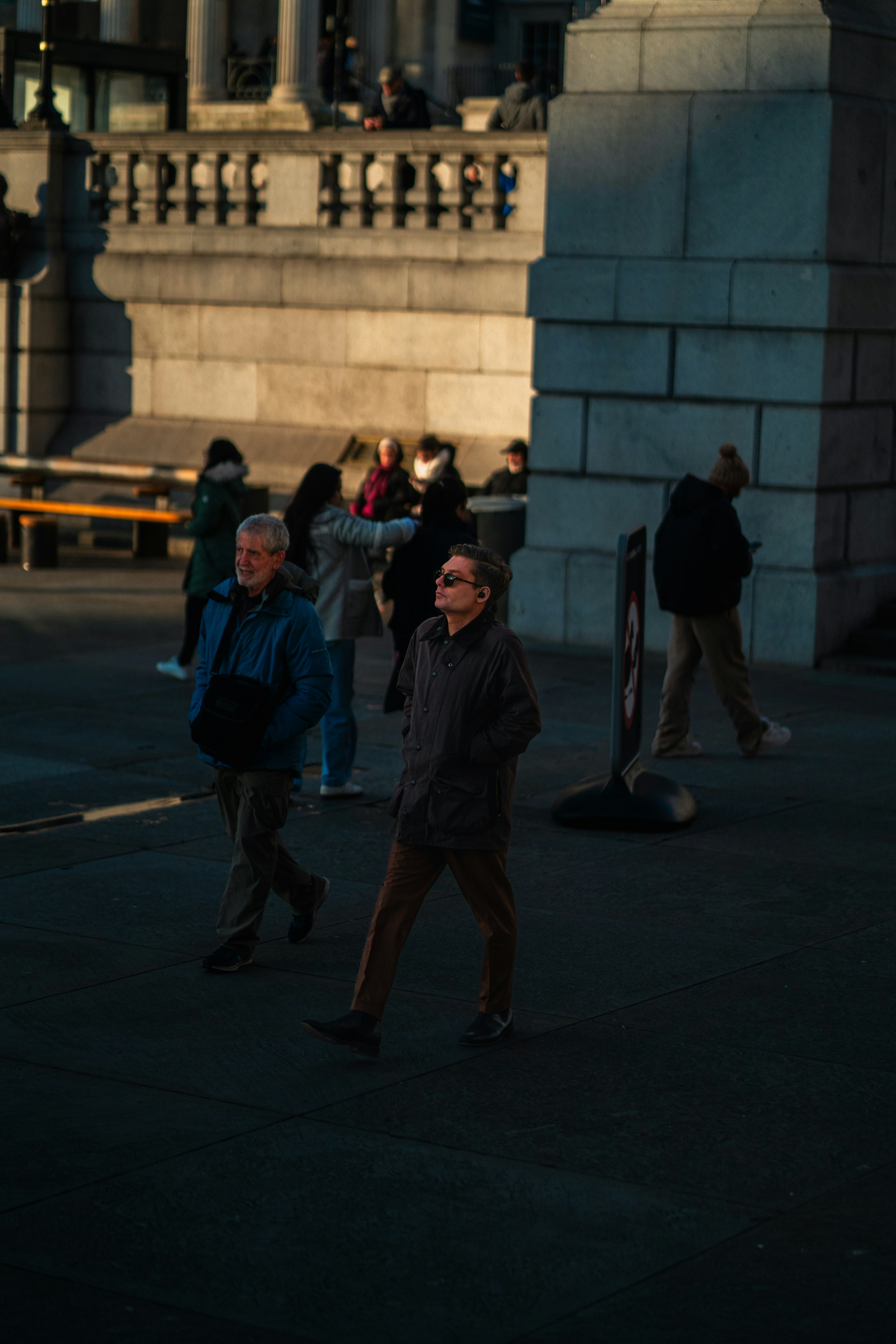Pedestrians stroll through a sunlit urban square with a grand stone building backdrop and a lone passerby in the foreground, captured in muted evening tones. This is a candid street scene photograph.