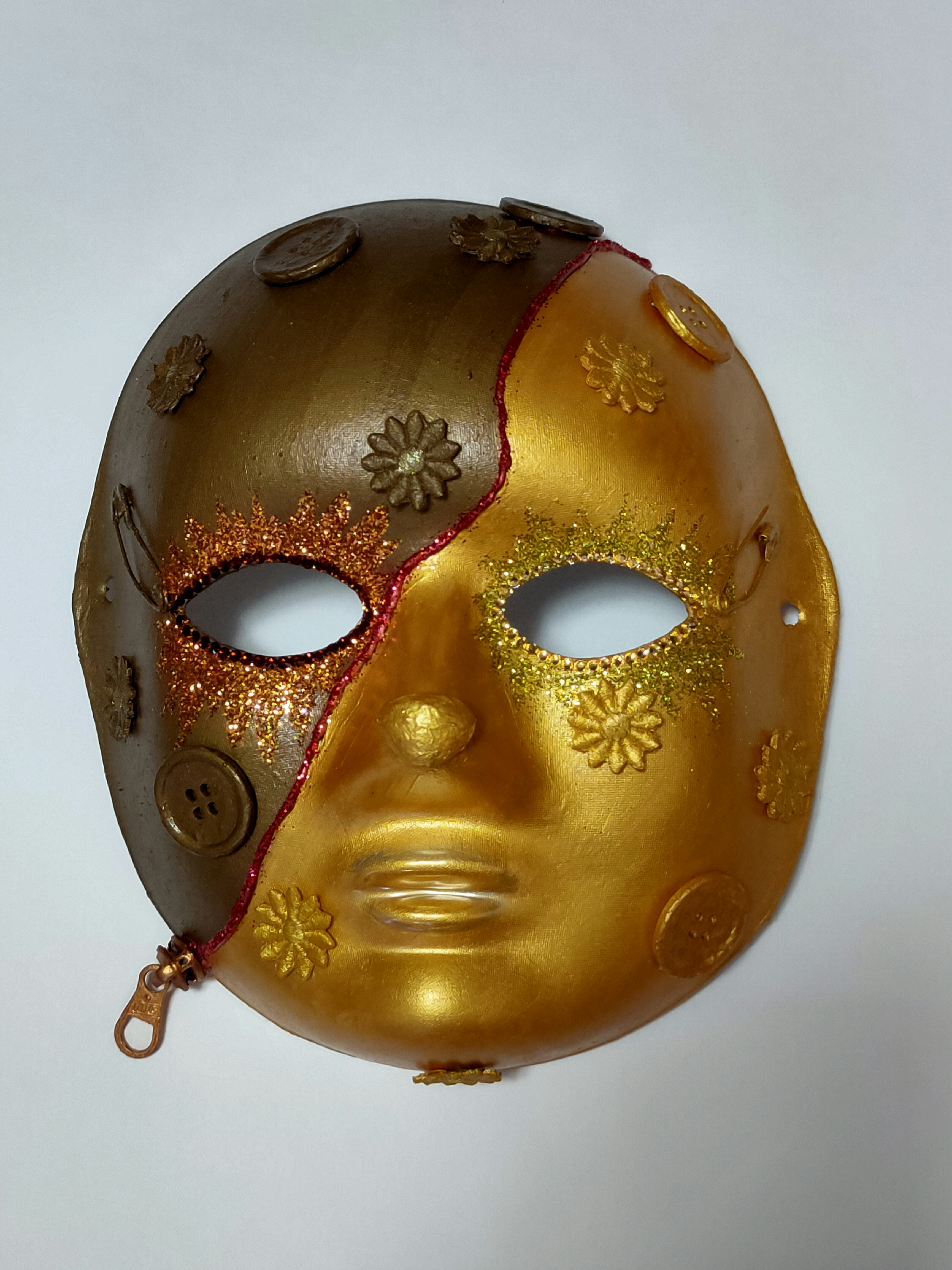 Intricately designed mask featuring a blend of gold and bronze hues adorned with floral patterns and buttons. The contrasting sides symbolize duality in expression.