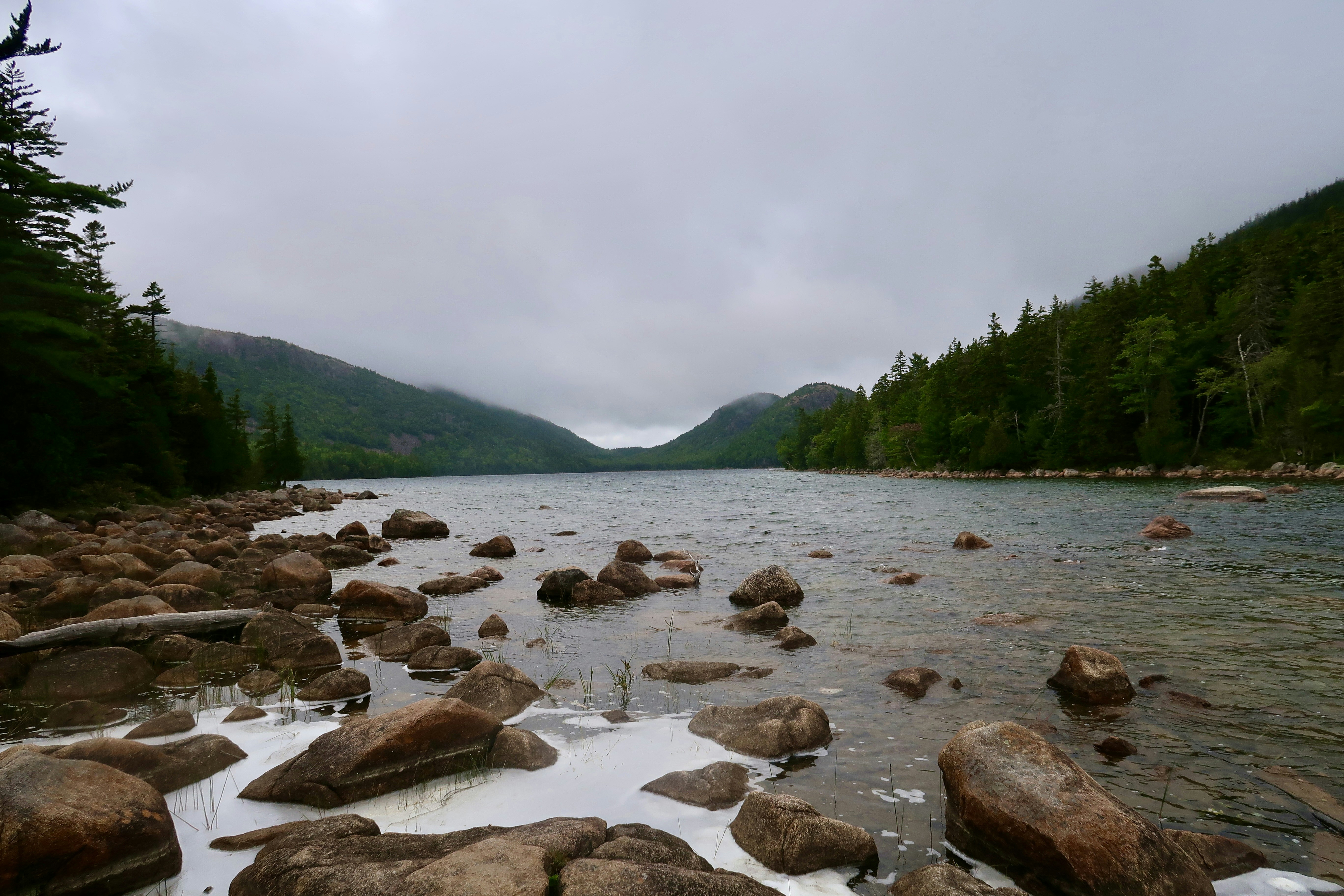Rocky shoreline of Jordan Pond under a cloudy sky with distant green hills.