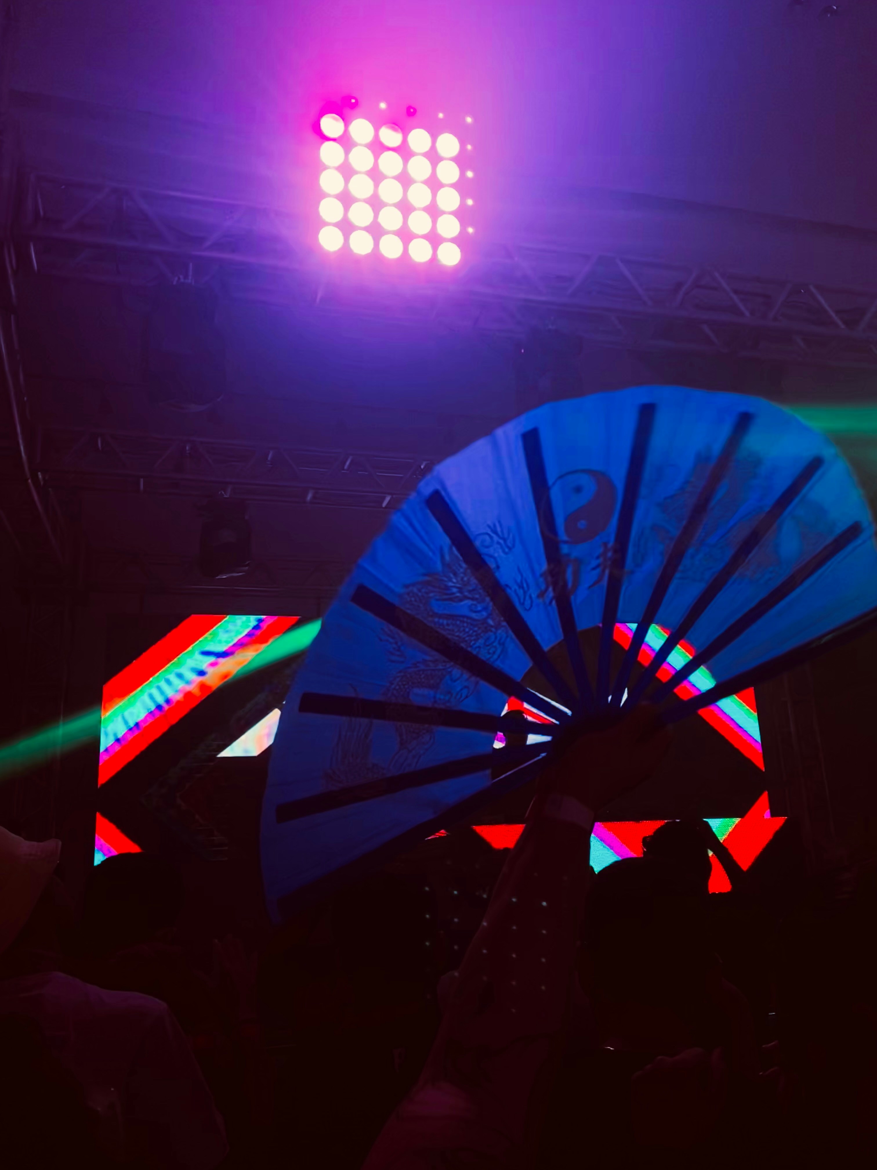A colorful fan held aloft against a backdrop of dynamic stage lights and visuals at a lively event. The scene captures the fusion of tradition and modern entertainment.