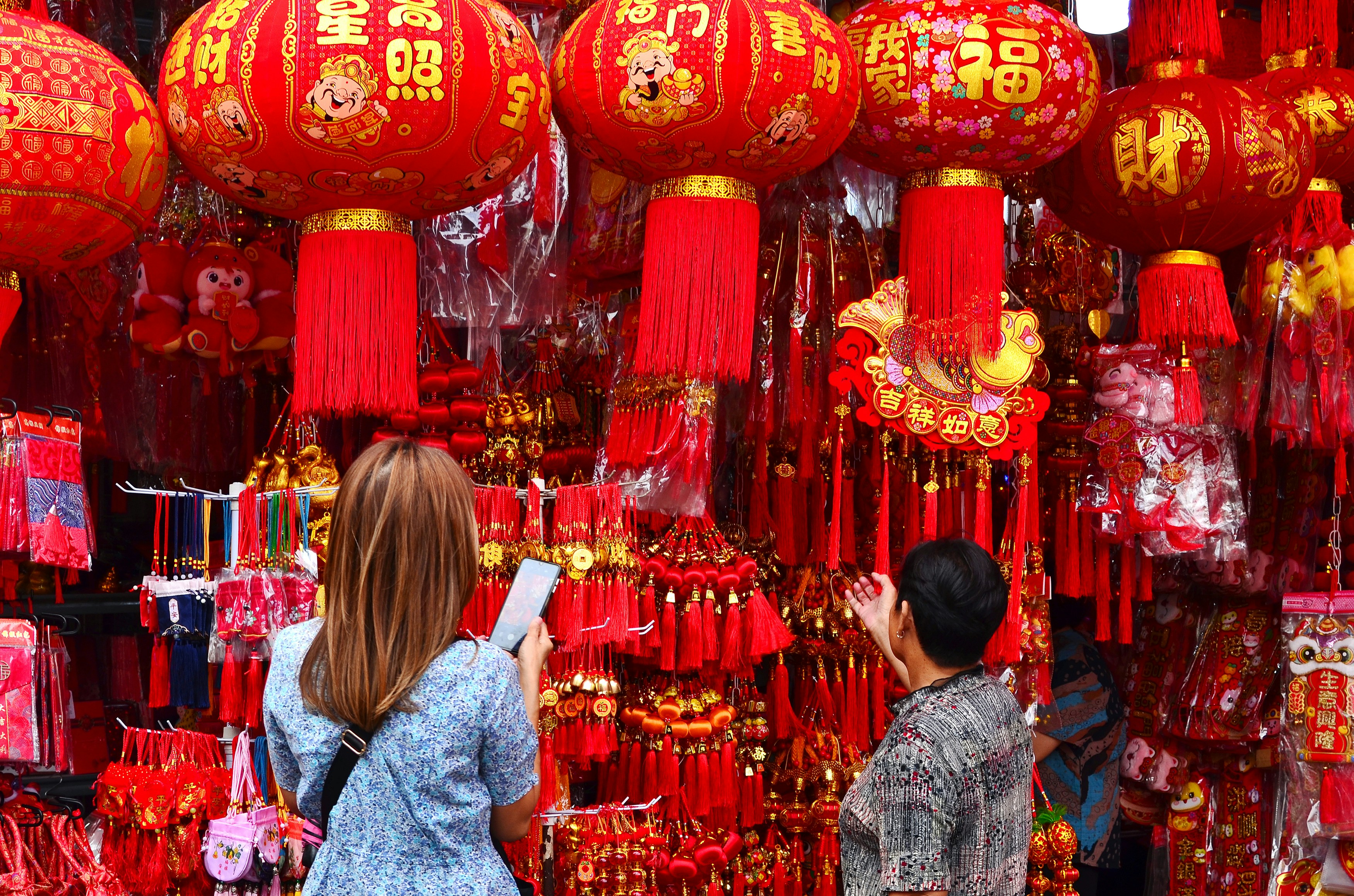 Two people browsing red and gold Lunar New Year decorations at a bustling market stall.