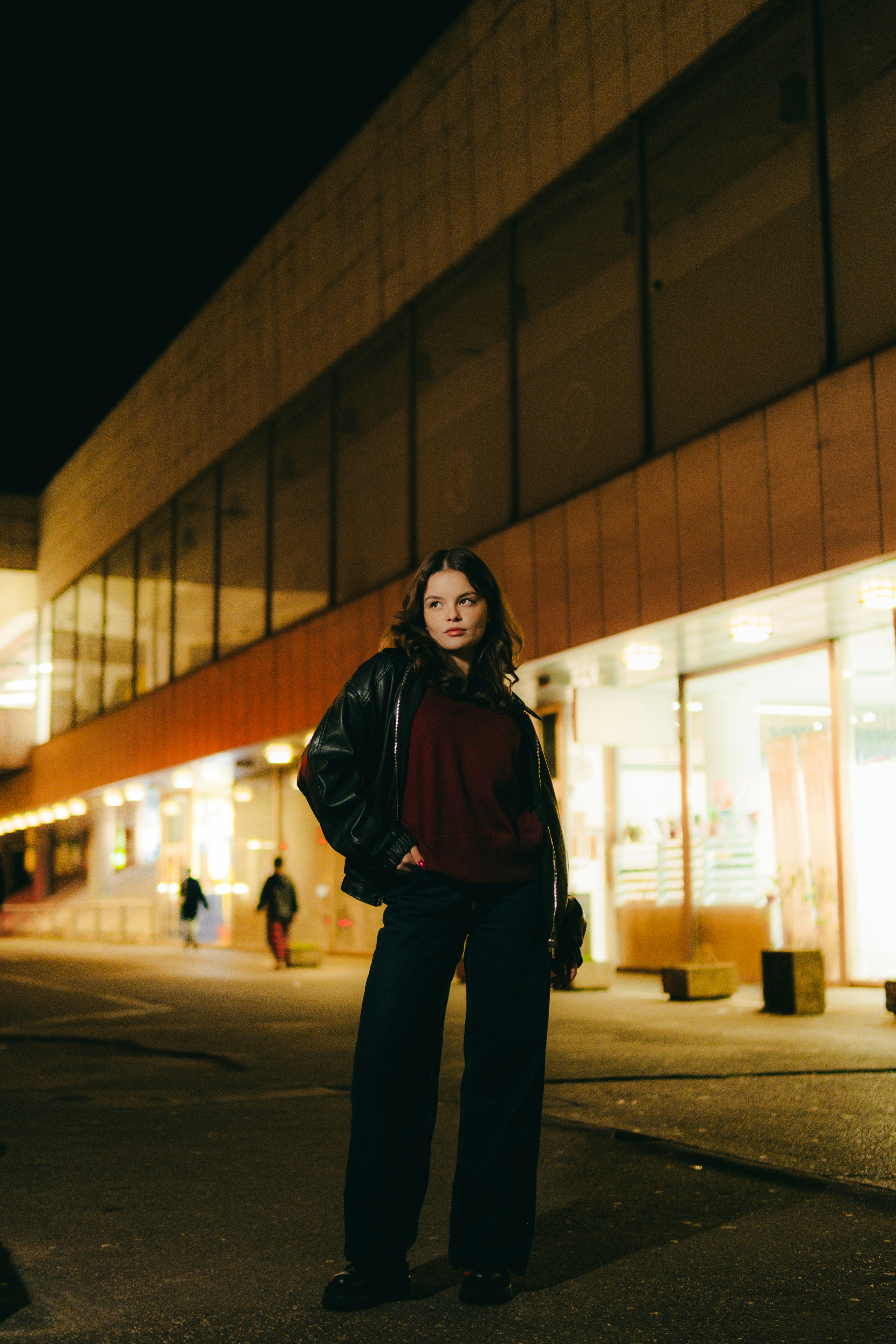 Woman in a dark leather jacket stands confidently on a dimly lit street, framed by the warm glow of city lights and ambient building illumination.