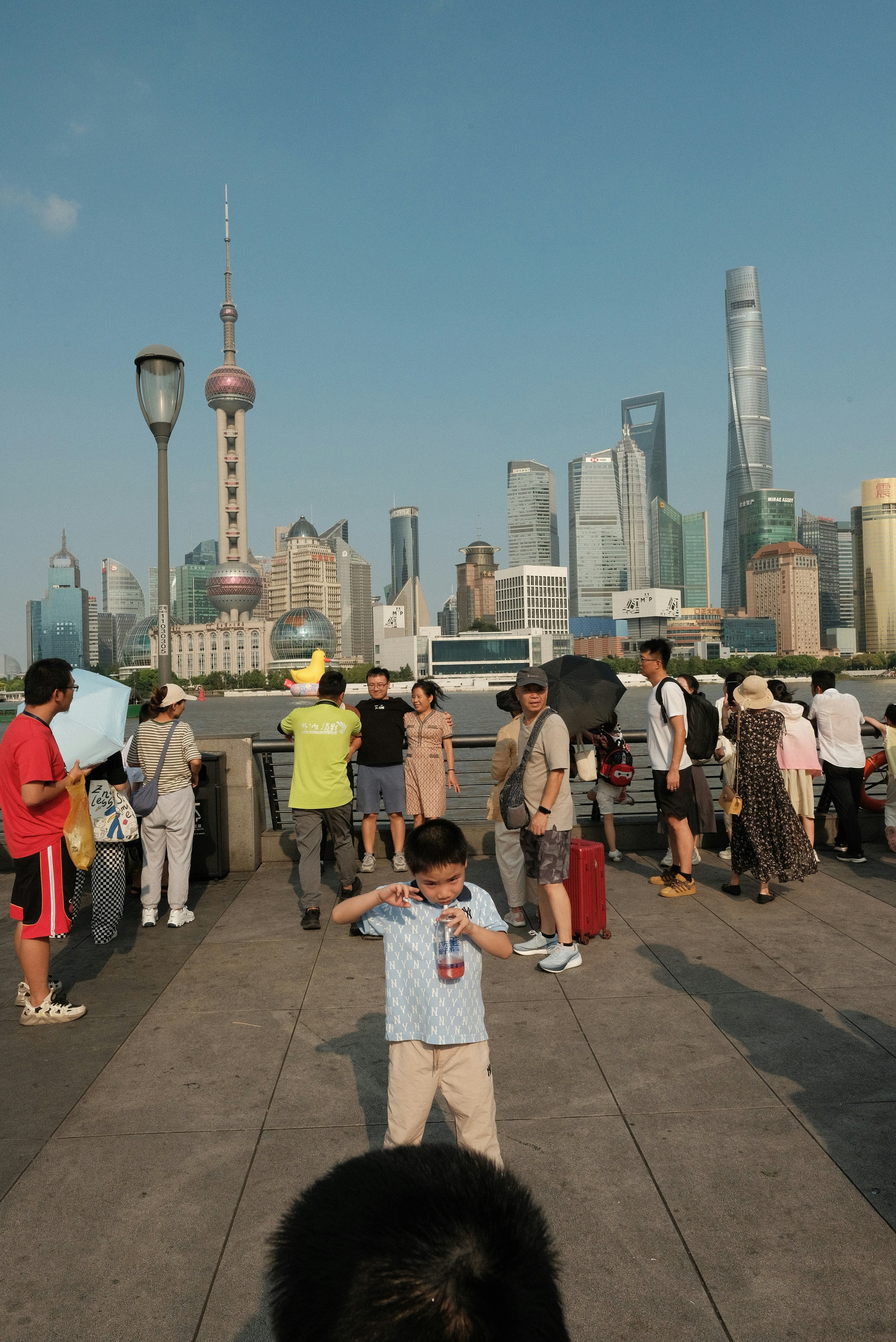 Crowd gathered on a riverfront promenade with Shanghai's skyline in the background under a clear blue sky.