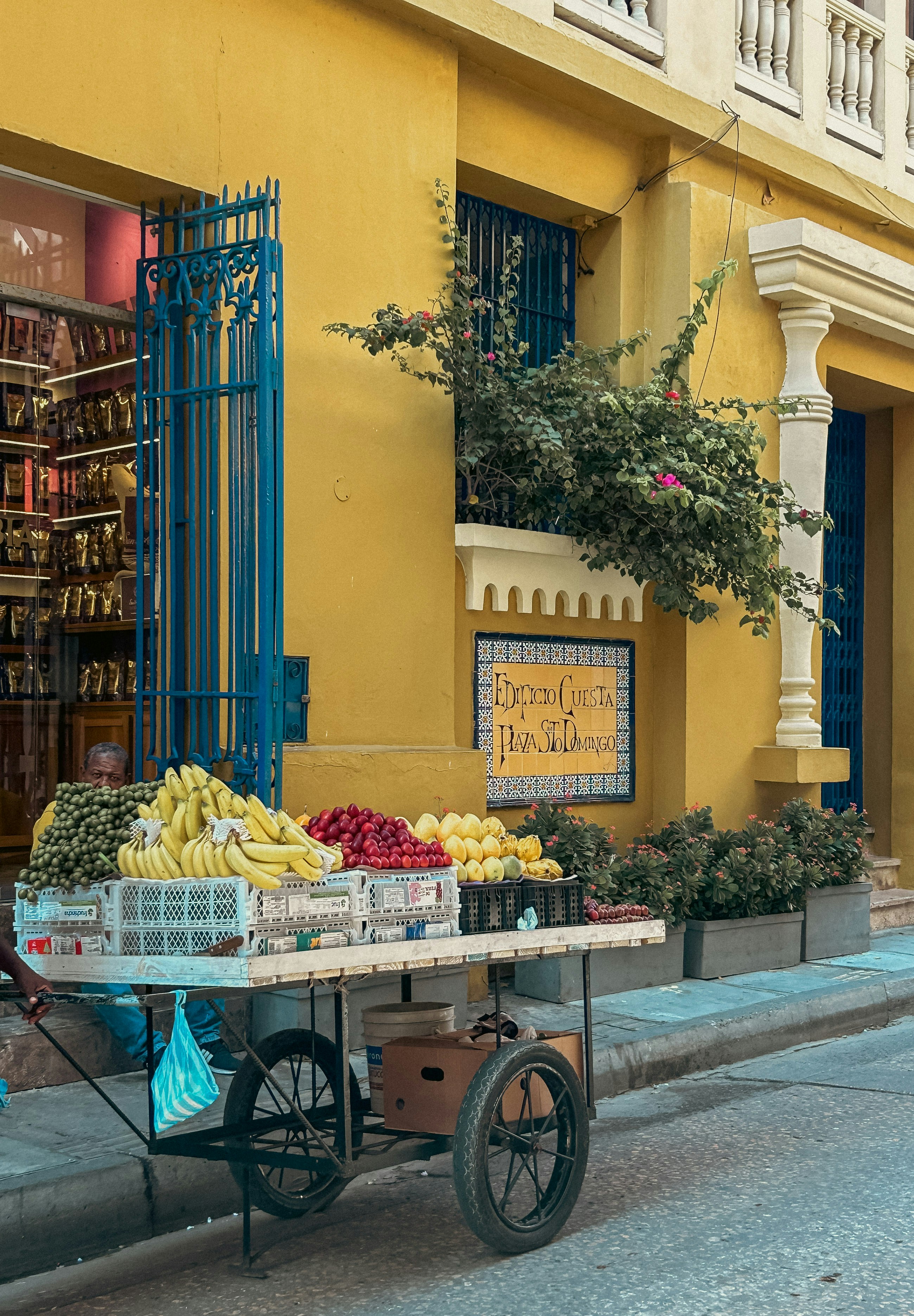 Fruit cart with bananas and lemons set against a bright yellow building with blue accents.