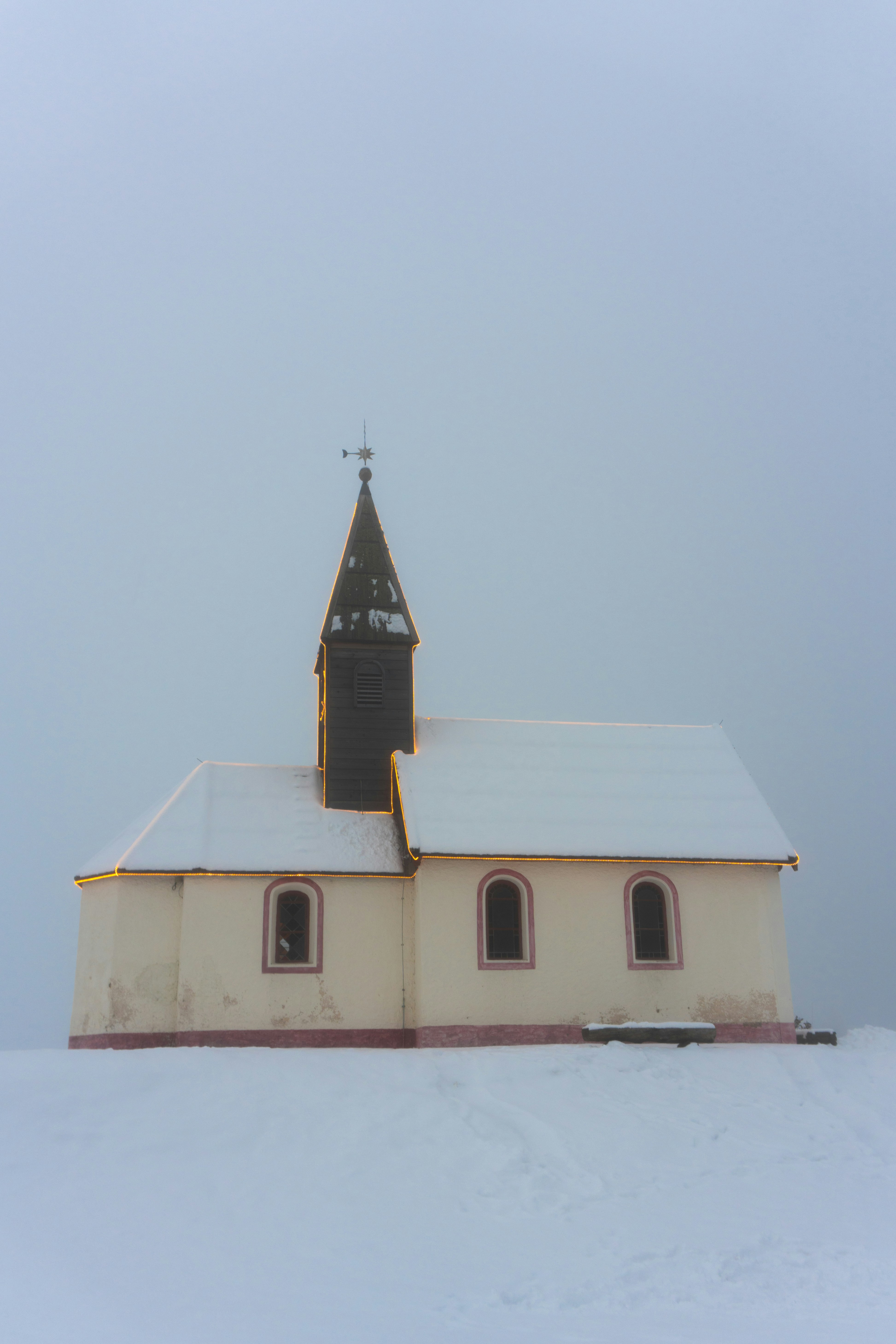 A snow-covered chapel with a dark steeple and warm edge lighting along the roof, set against a pale blue winter sky.