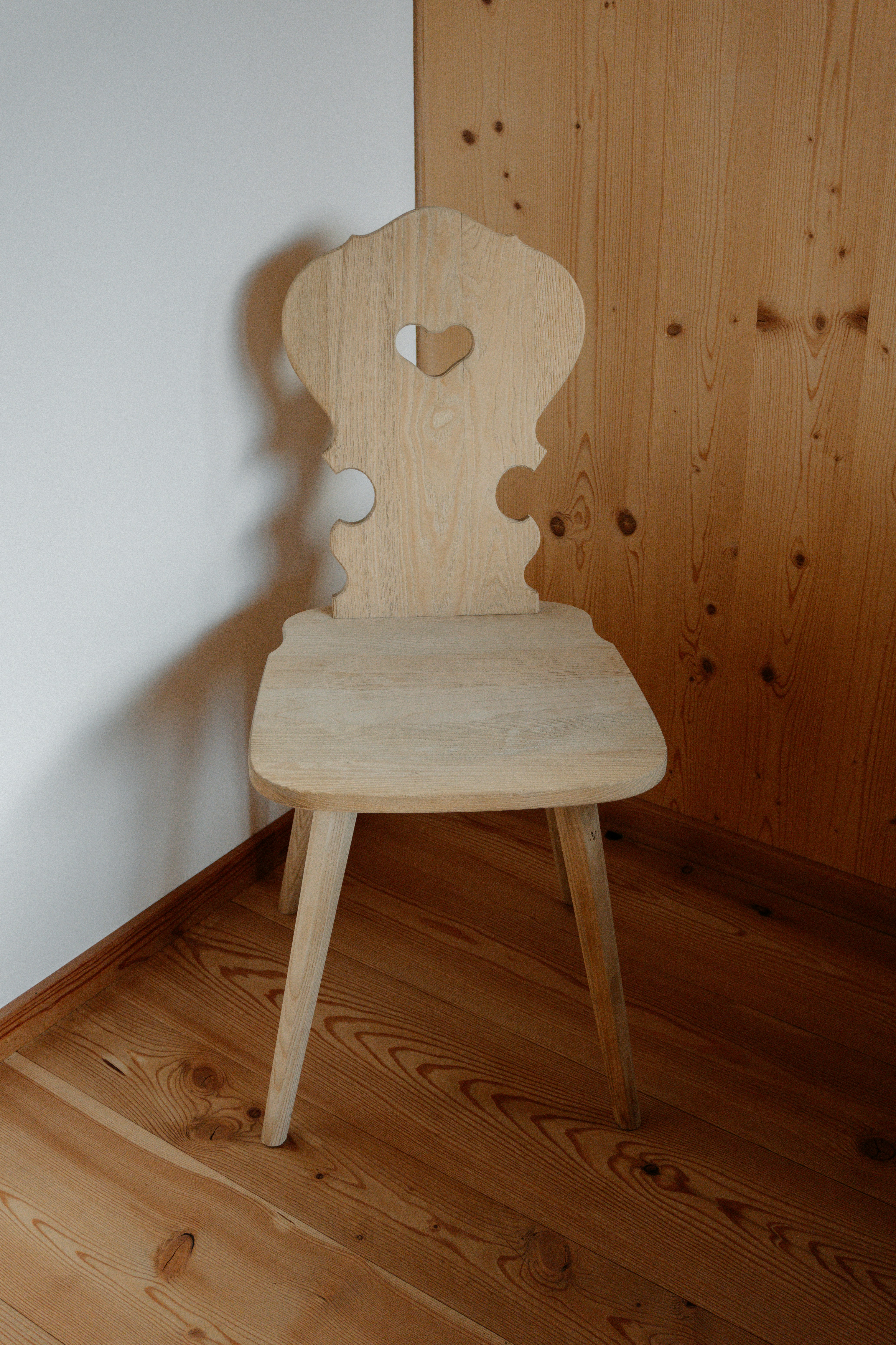 Light wooden chair with a heart-shaped cutout in the backrest sits in a corner of a wood-paneled room.