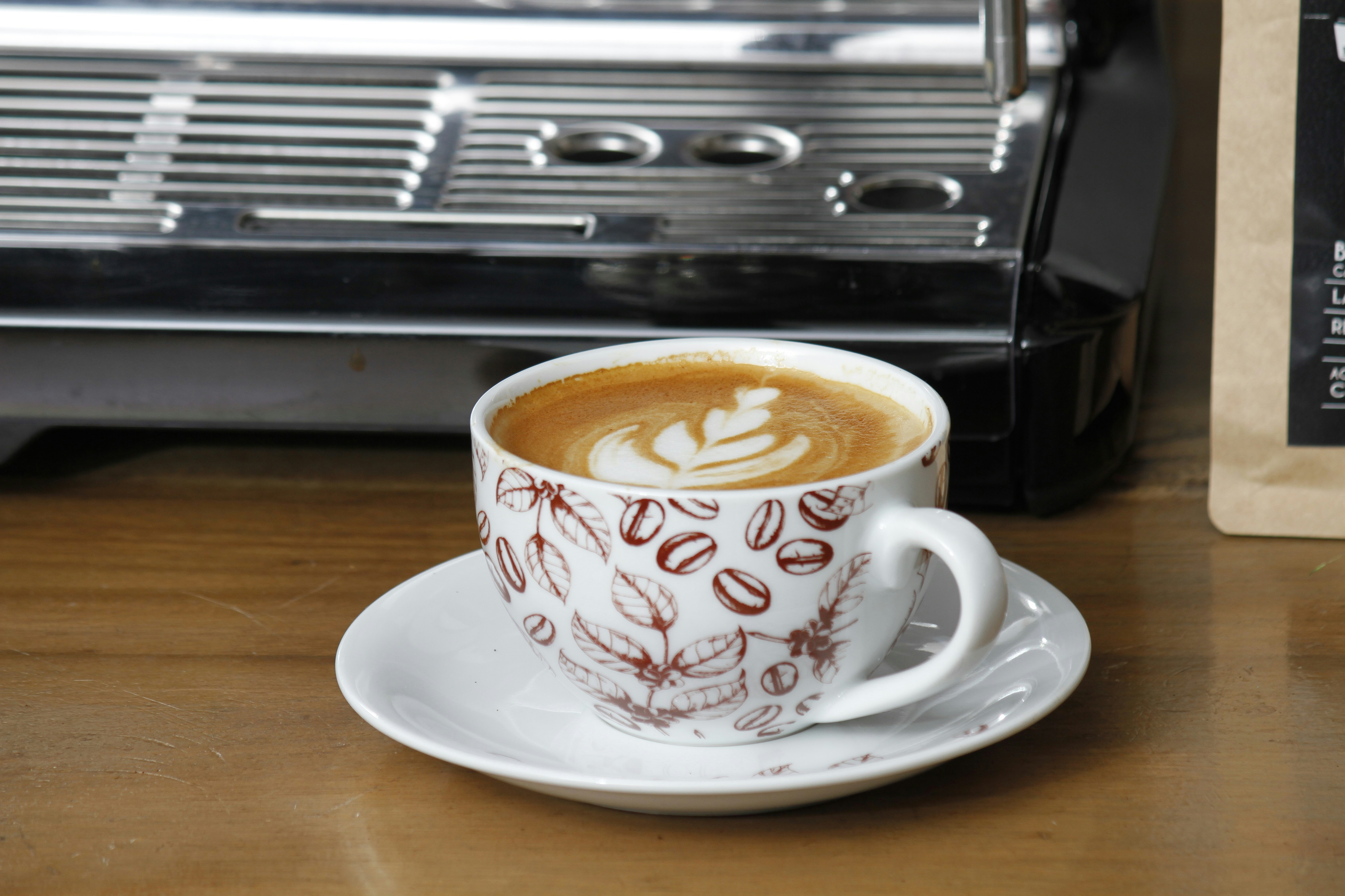 Latte art in a patterned cup beside a sleek espresso machine on a wooden counter.