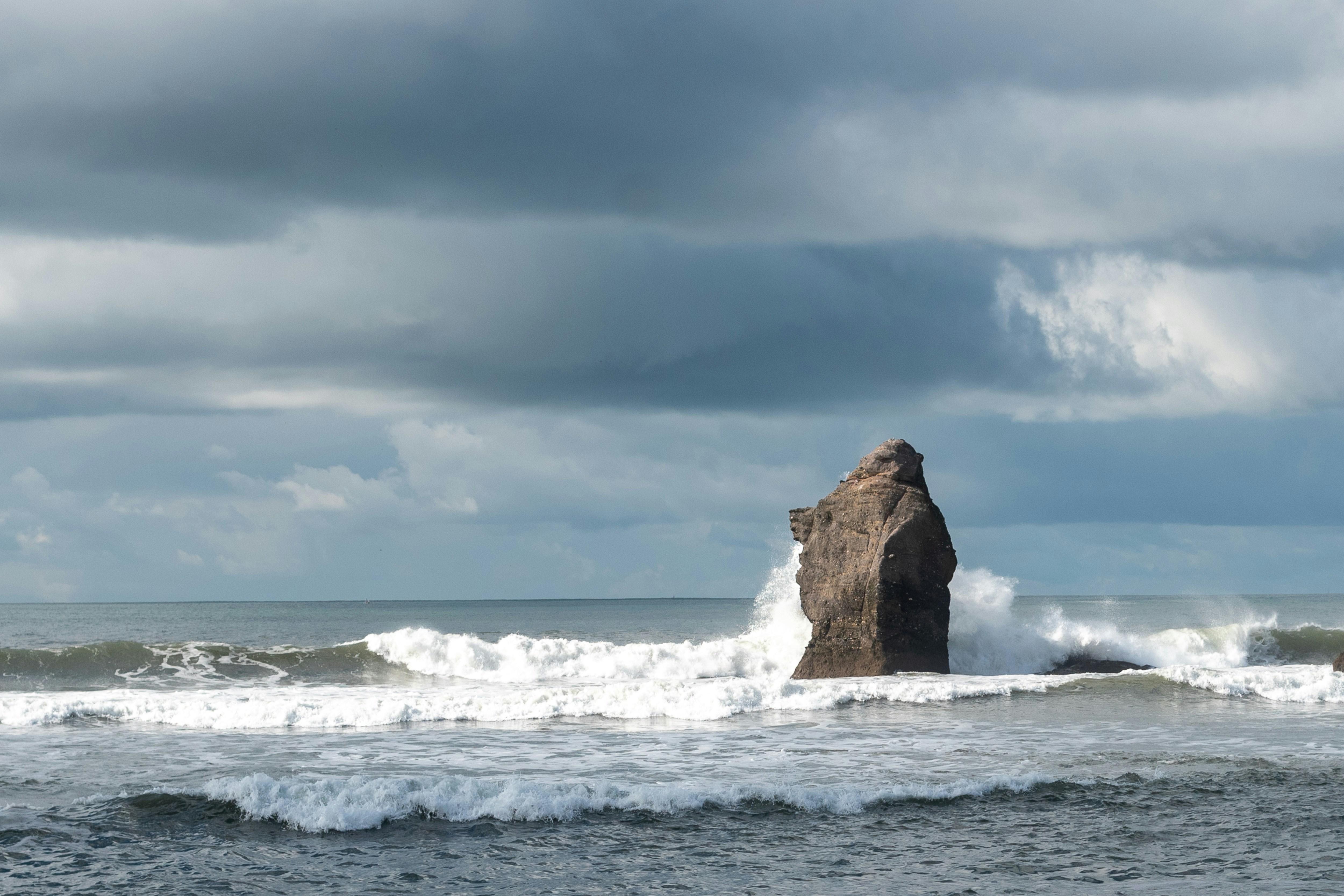 Ocean waves crash against a lone rock formation under a dramatic cloudy sky.
