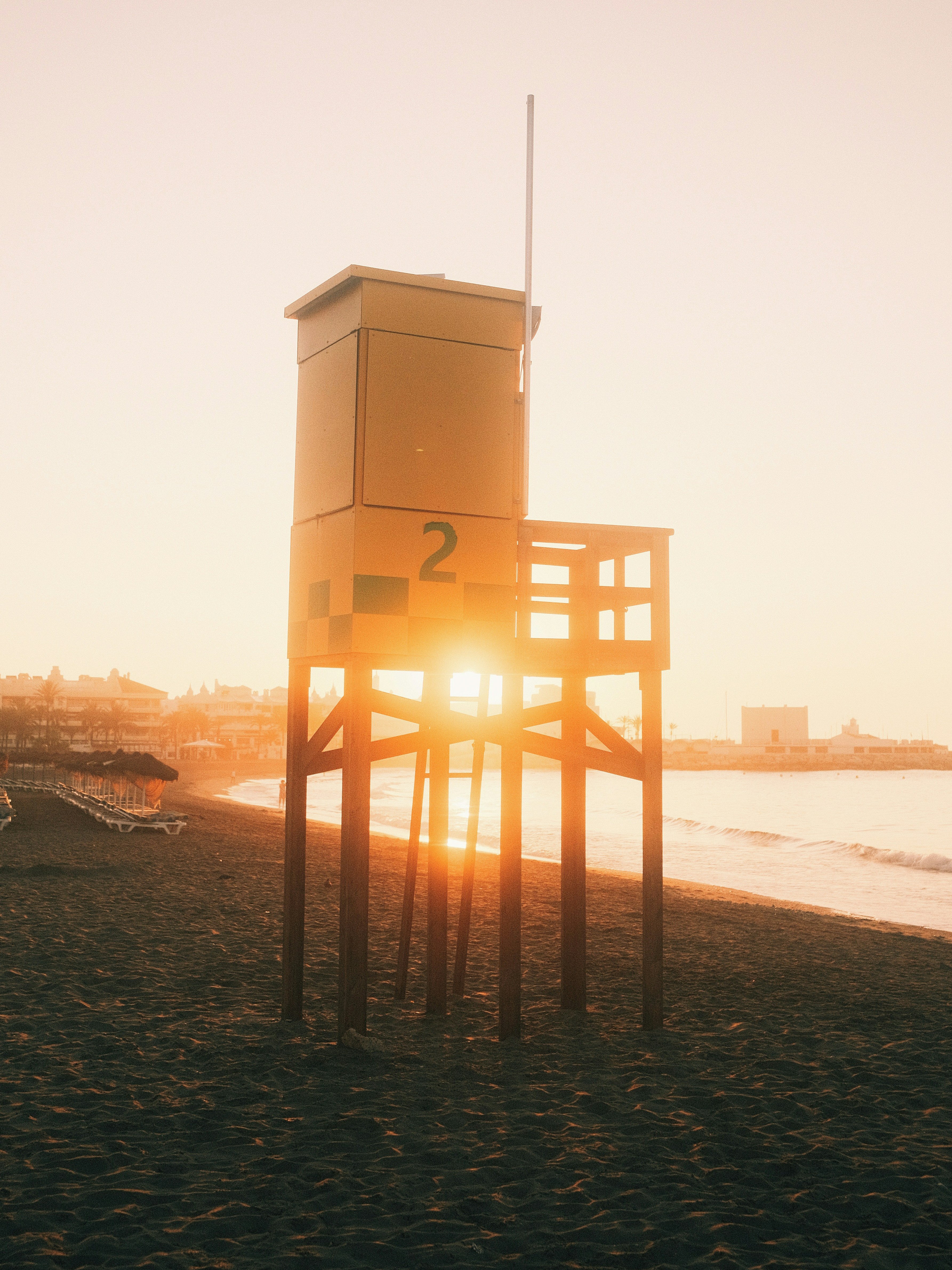 Lifeguard tower silhouetted against a warm sunset on a sandy beach, with the sun peeking through its legs casting long shadows.