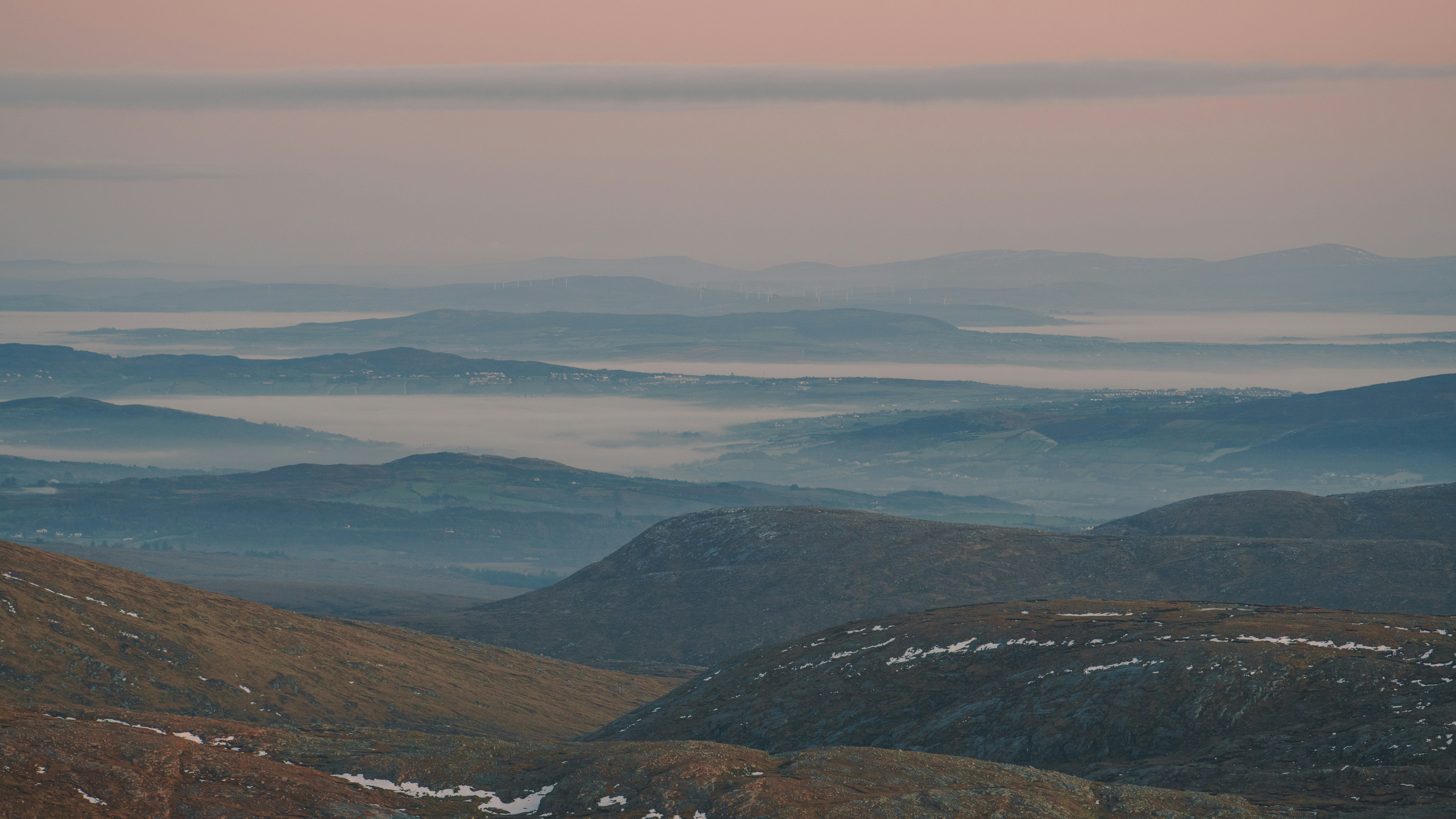 Rolling hills under a soft pink sky with layers of mist settling between them.