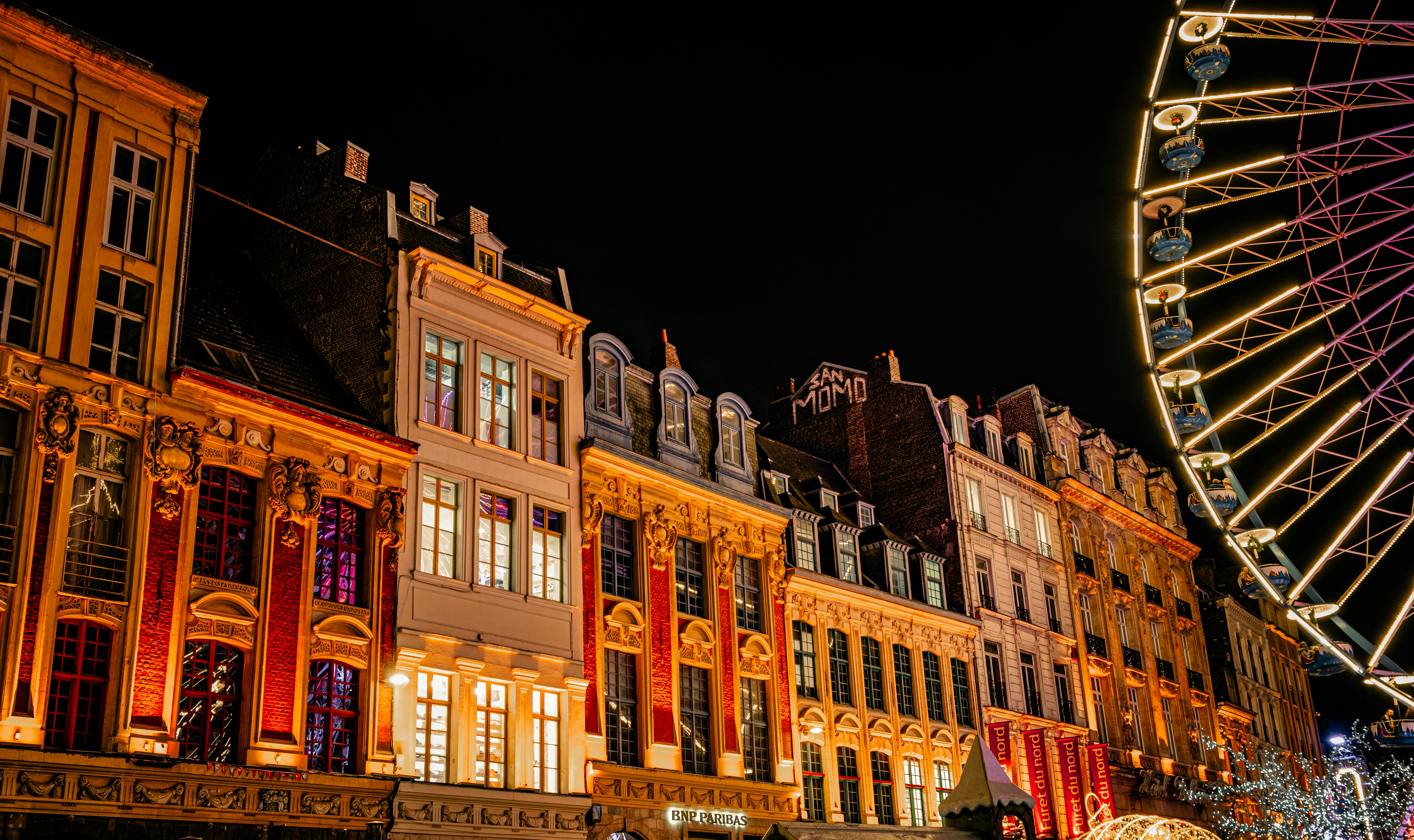 Illuminated historic buildings in Lille with a Ferris wheel partially visible against the night sky.