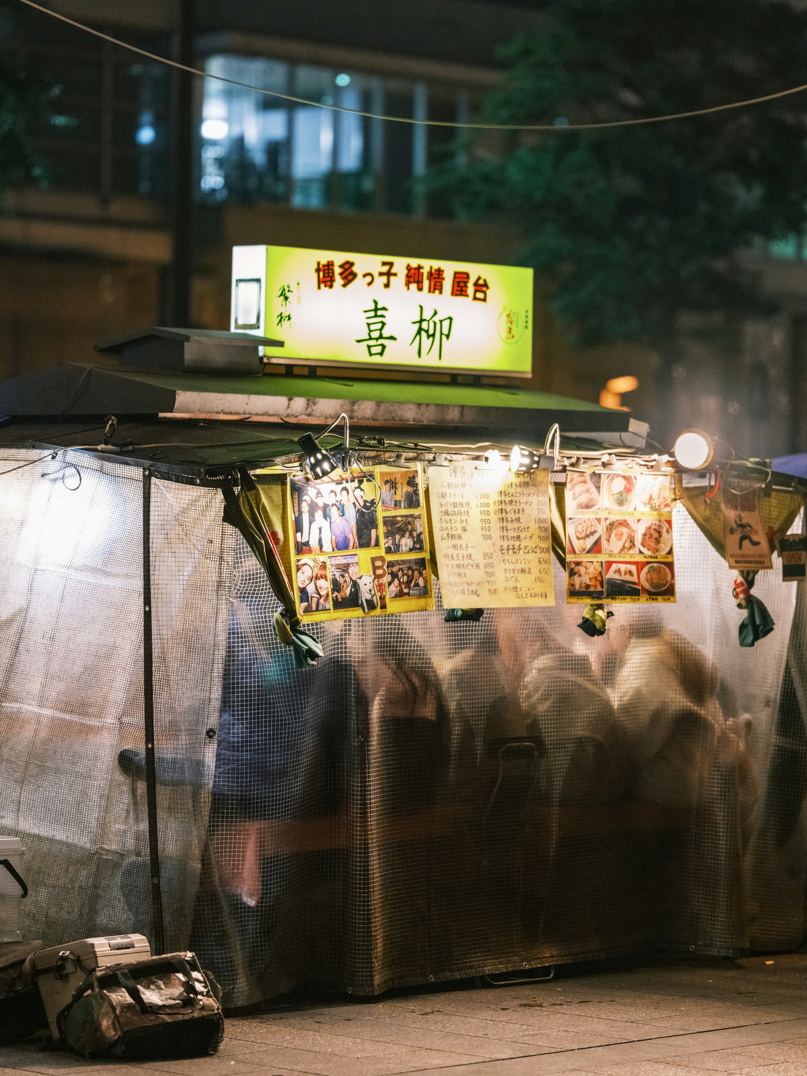 Photograph of a night market stall at night with a glowing sign and plastic-walled exterior. Blurred figures behind the coverings hint at activity.