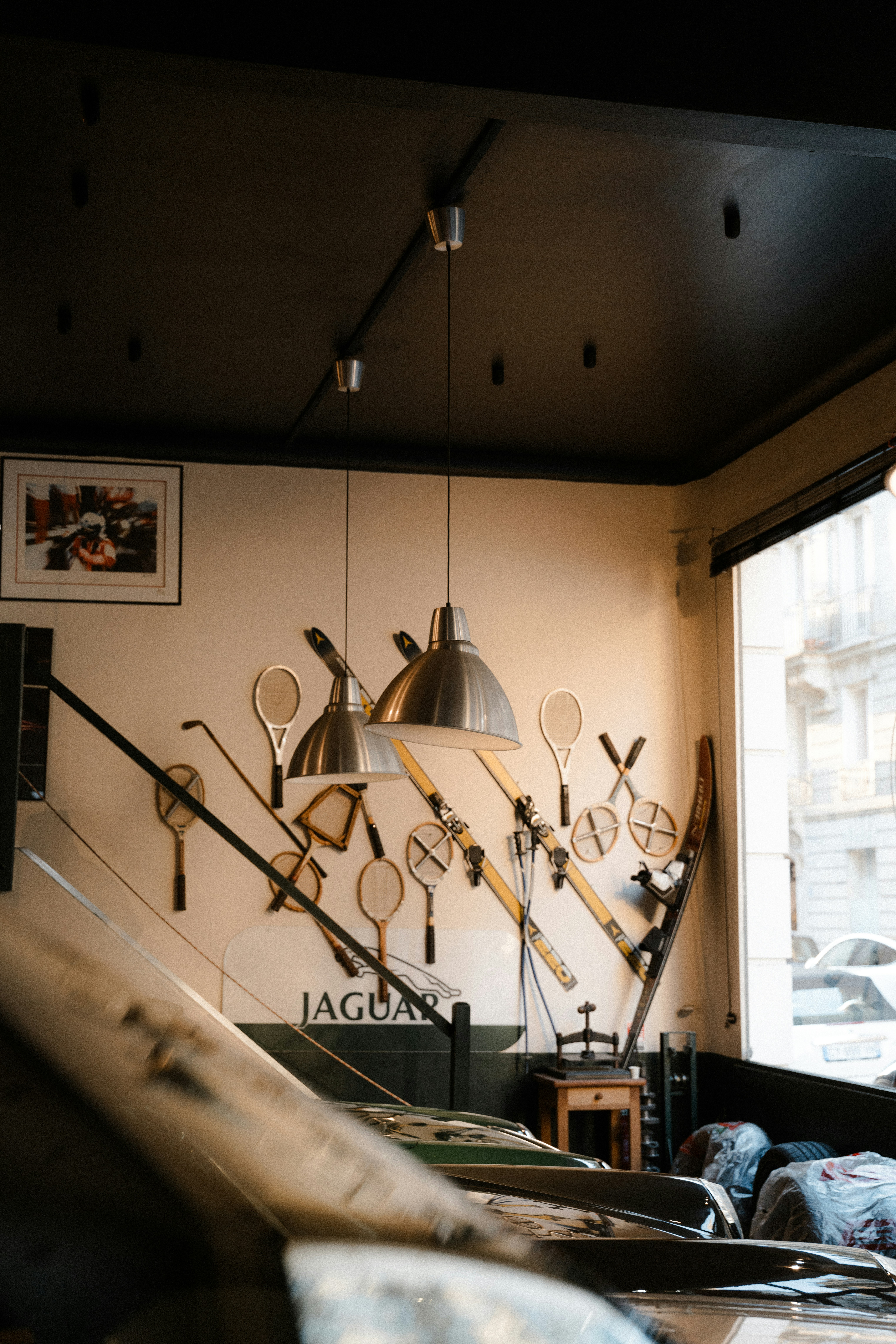 Vintage sports gear, including tennis rackets and skis, hangs on a cream wall under metallic pendant lights, creating a warm, nostalgic ambiance.