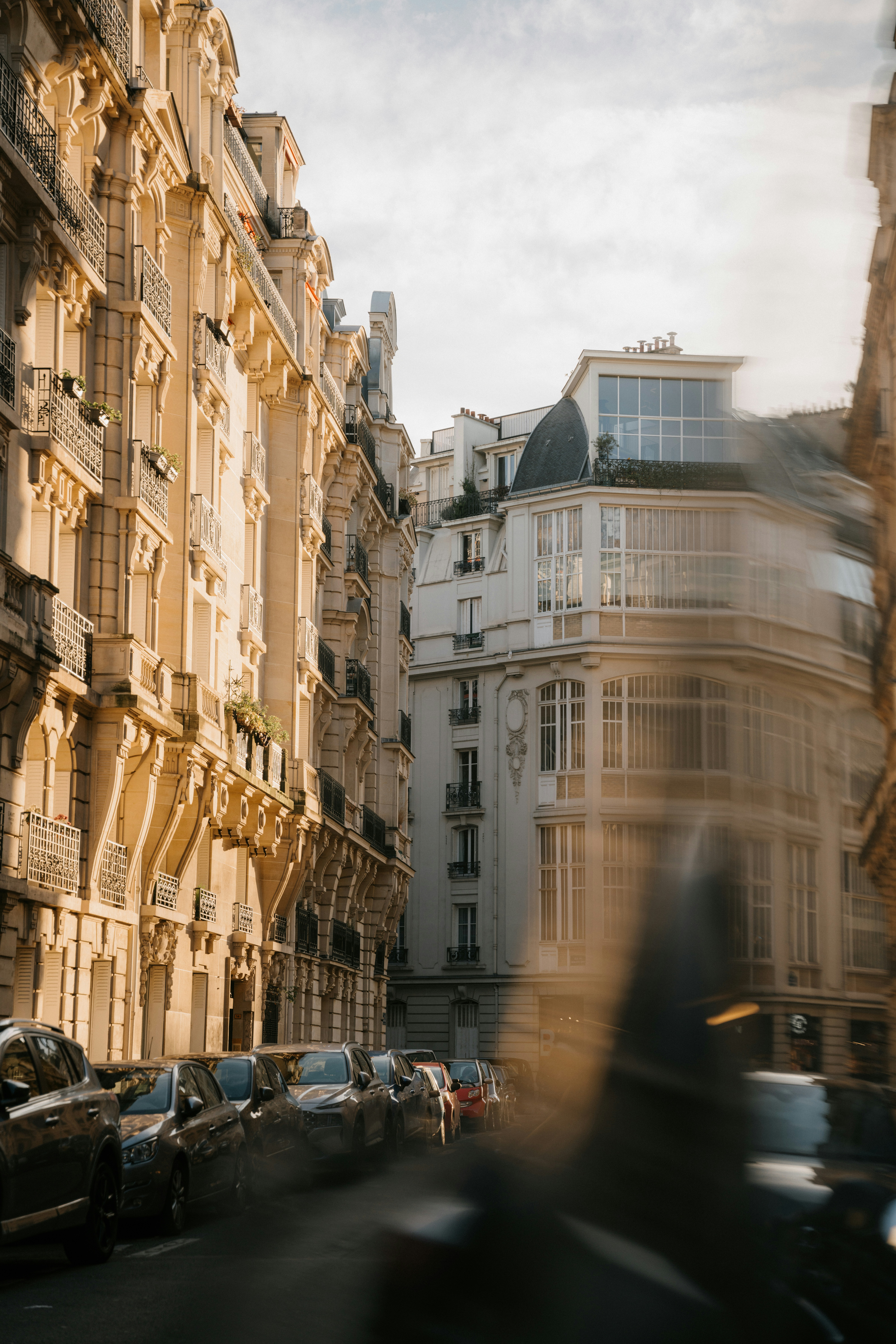 Blurred figure moves through a sunlit Paris street flanked by ornate buildings.