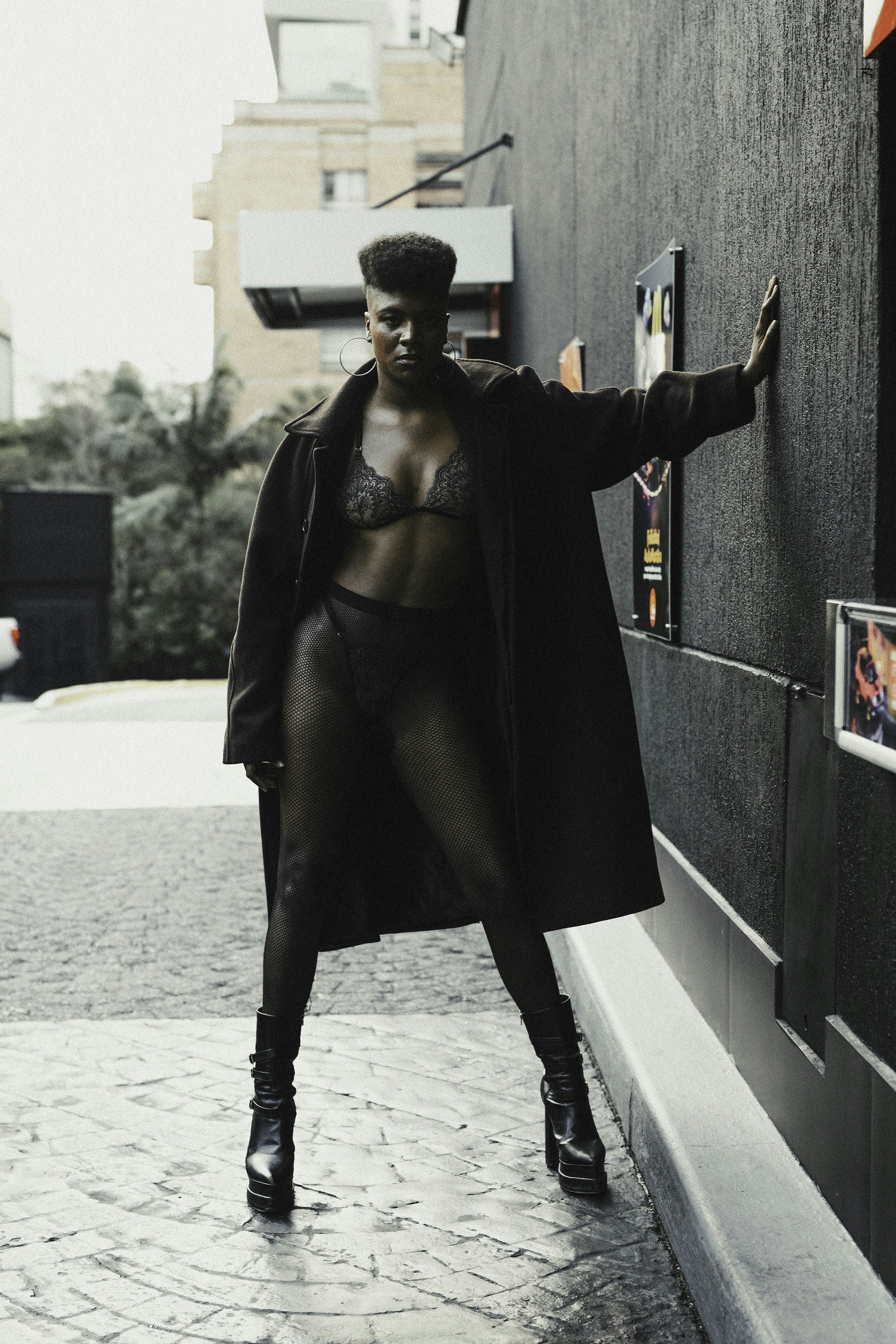 Model in black lingerie and a long coat poses in a narrow urban alley, leaning against a dark wall. Gritty city backdrop emphasizes the texture of the outfit and the mood of urban fashion.