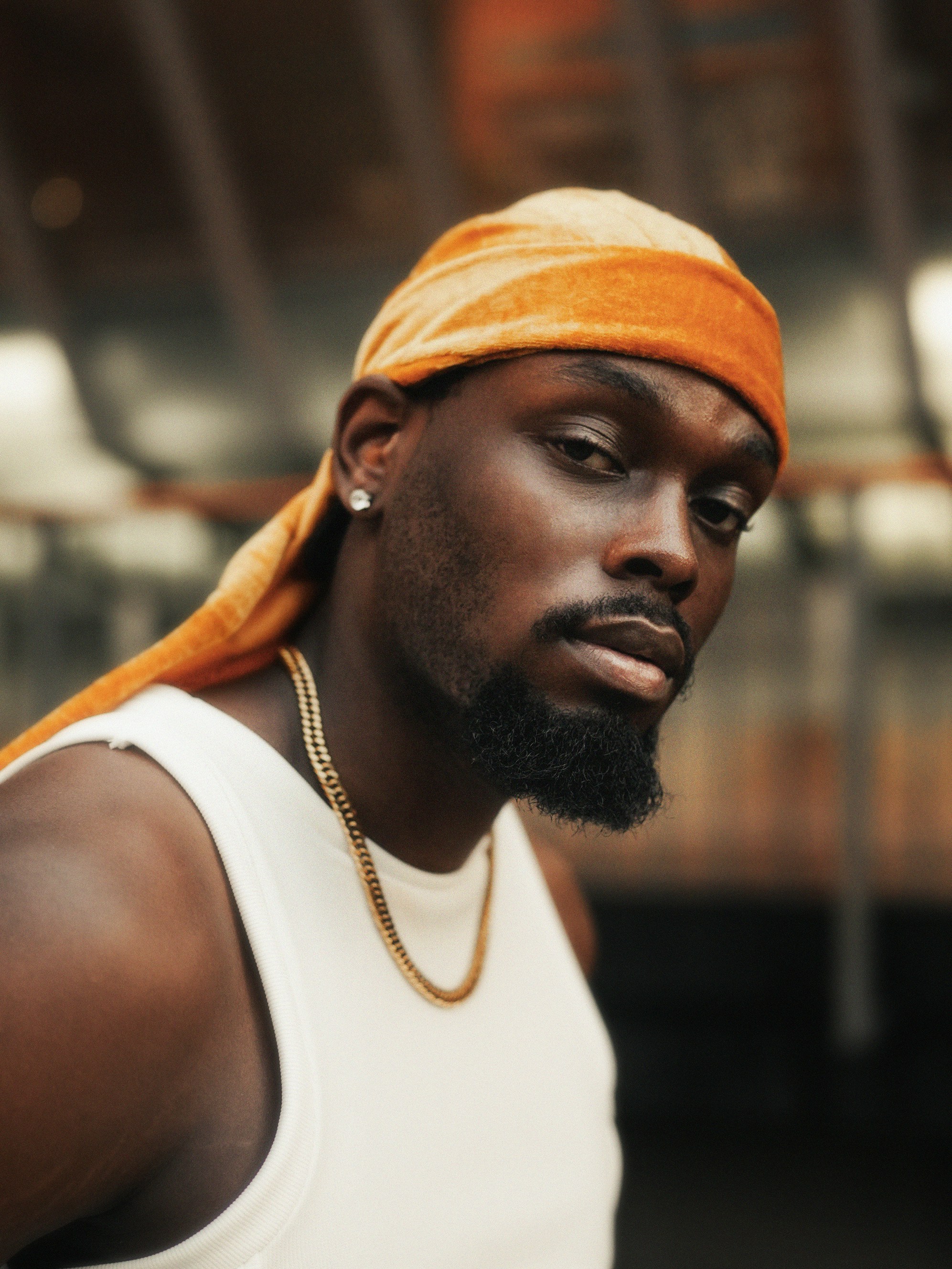 Man in an orange headscarf and white tank top against a softly blurred background. Warm lighting enhances skin tones and scarf color, highlighting an expressive gaze.