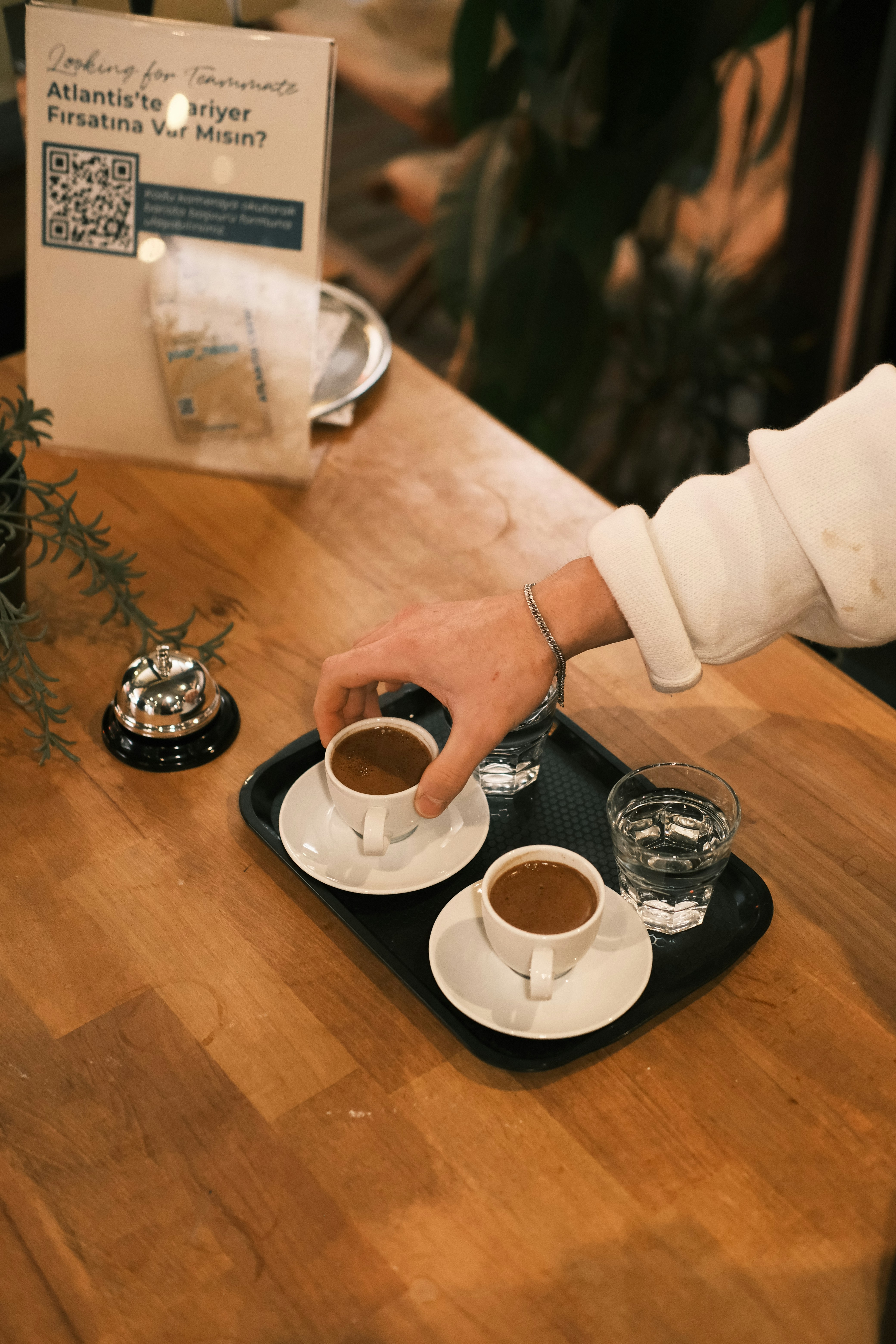 Photograph of a warm cafe counter with a hand reaching for two espresso cups on a black tray, beside a glass of water and a small bell. The soft amber lighting highlights the intimate moment and the wooden textures.
