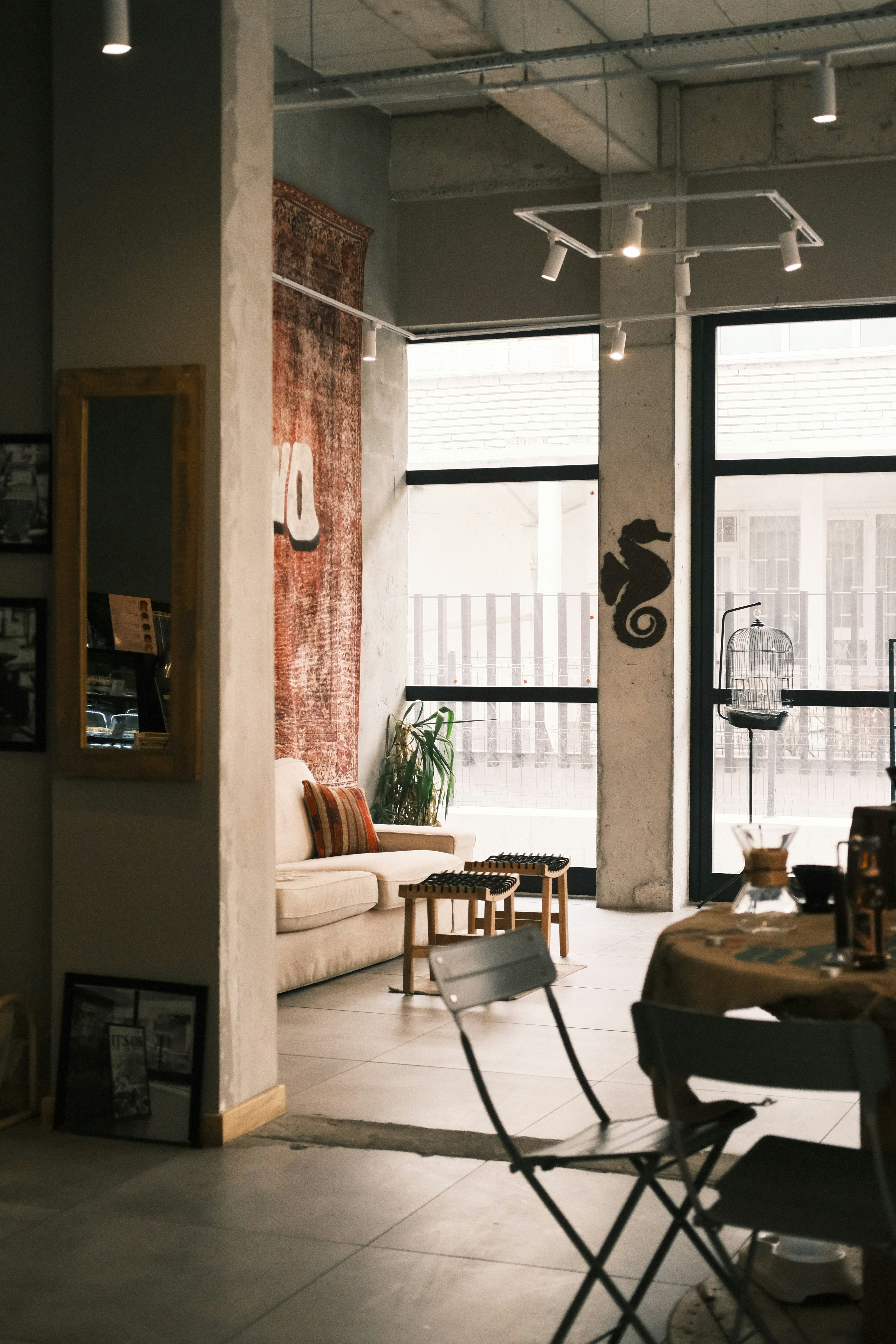 Warm coffee shop interior featuring a beige sofa, red tapestry, and seahorse wall art by large windows.