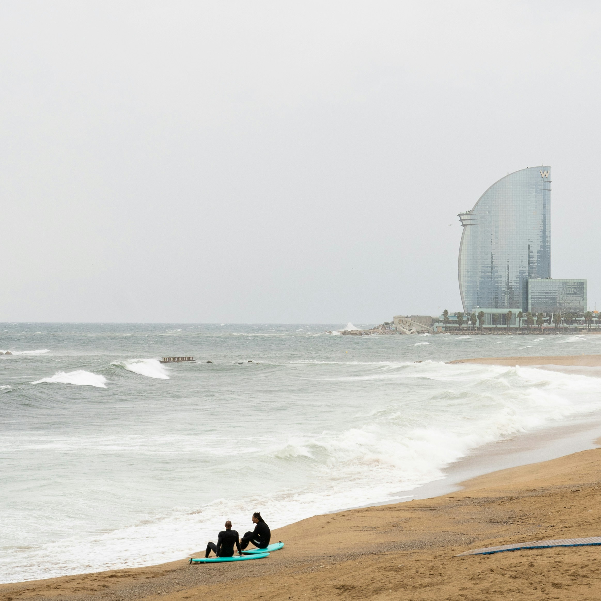Two Surfers on Quiet Shore as the W Barcelona Towers Over the Horizon