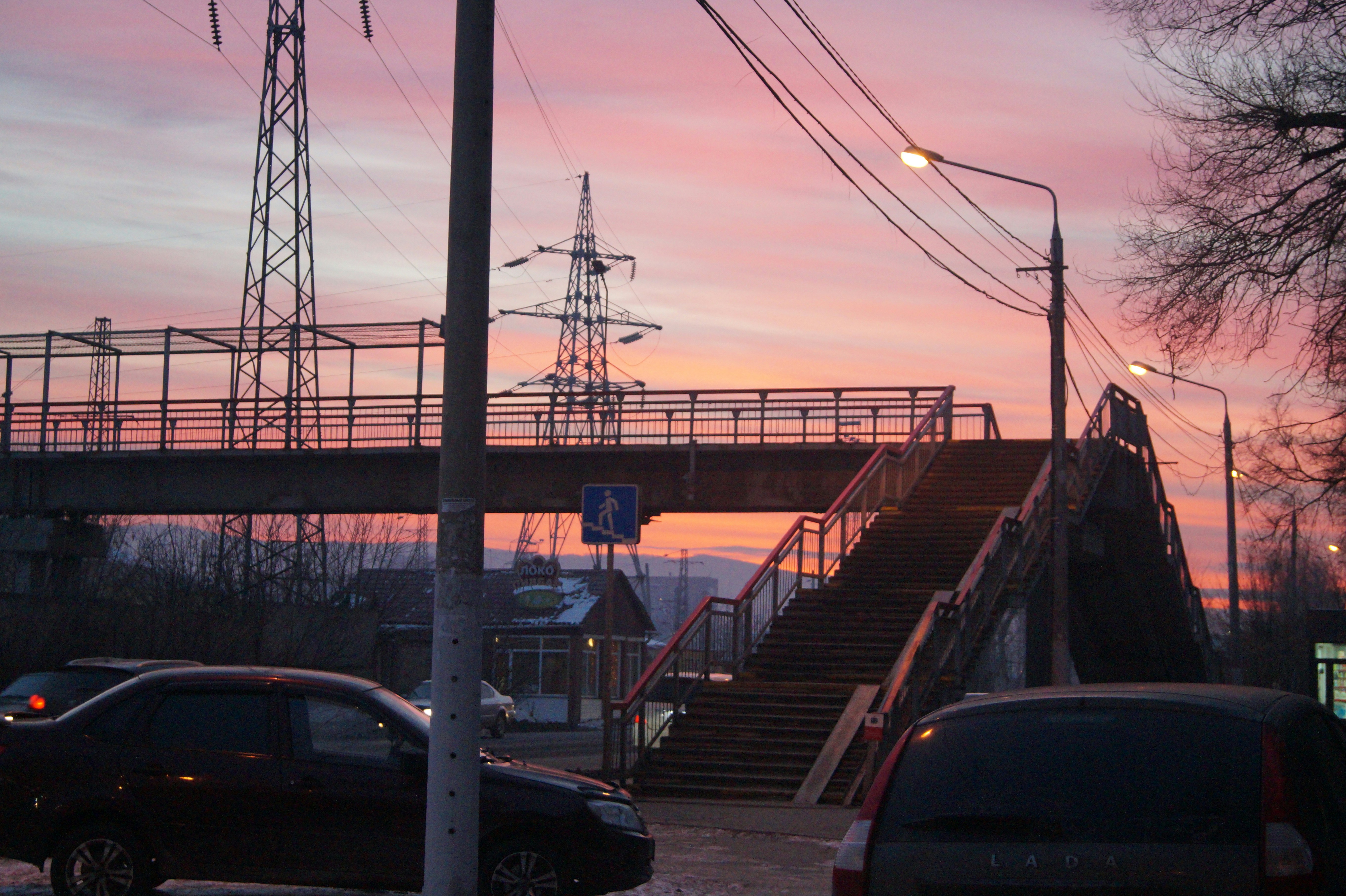 Dusk light over an urban overpass with stairway and parked cars, silhouetted by power lines and a soft pink-orange sky.