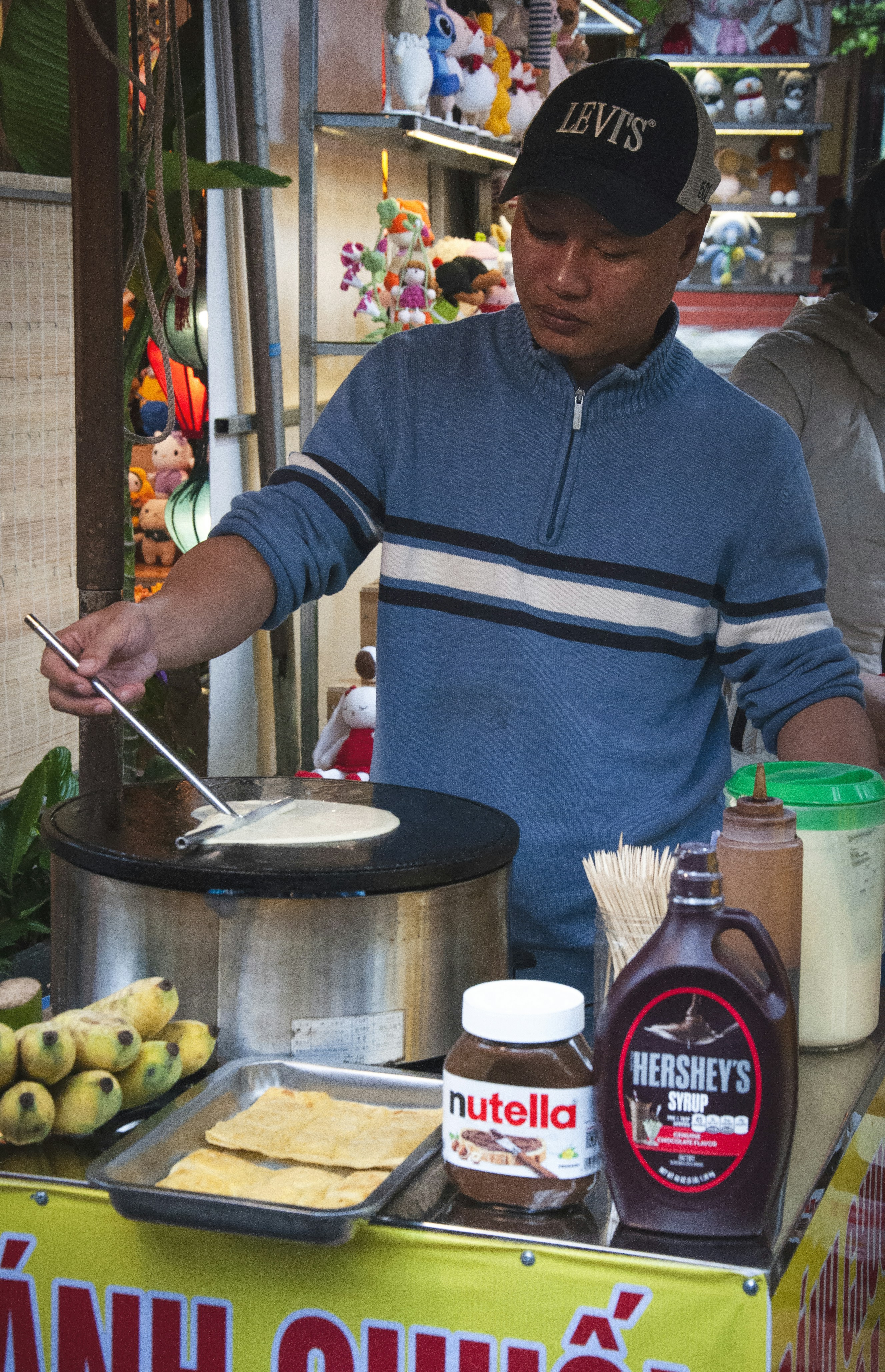Street vendor expertly preparing a crepe, surrounded by colorful plush toys and various toppings. A blend of culinary art and vibrant market life.