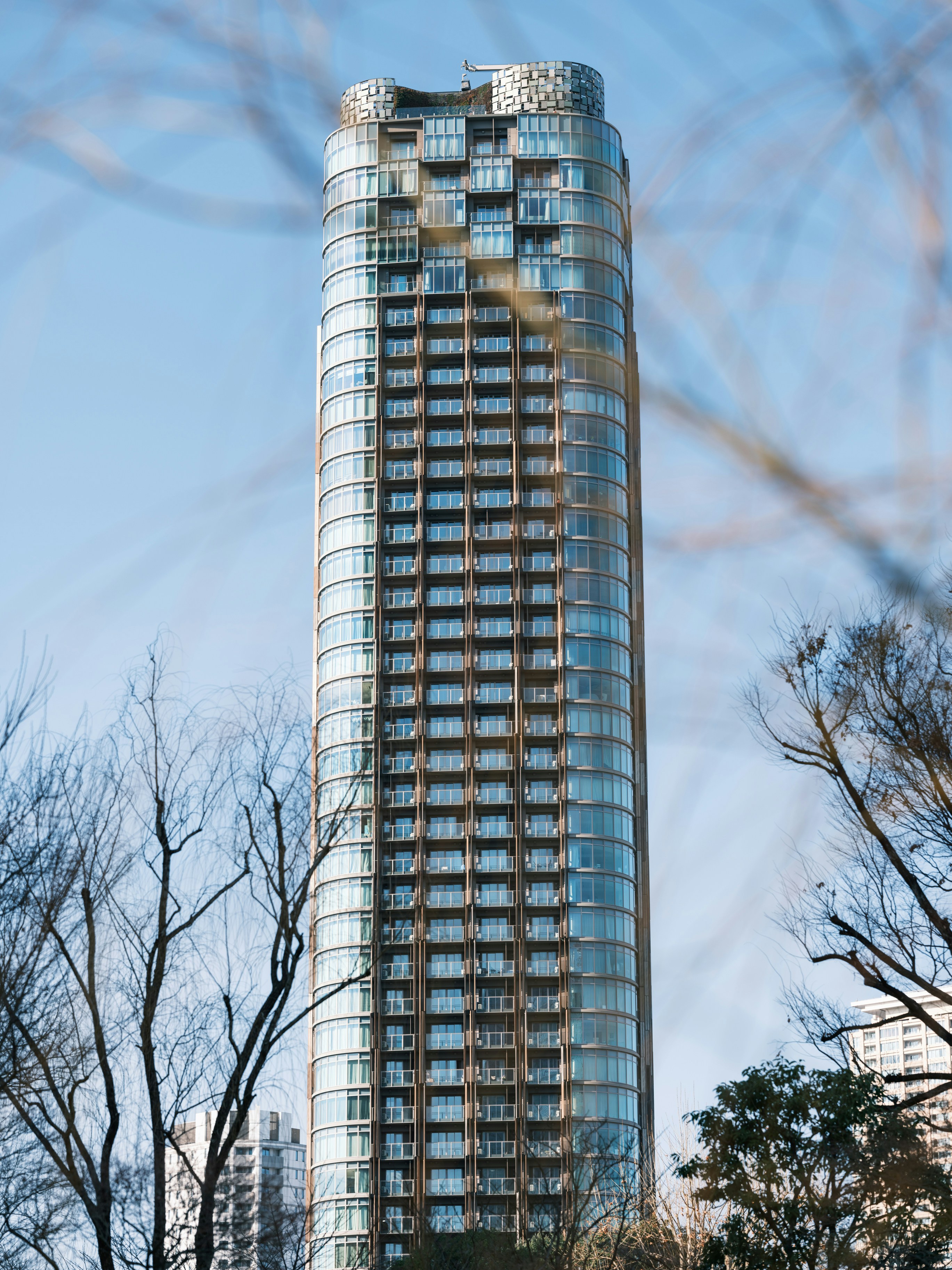 Tall cylindrical glass-clad skyscraper rises above bare trees against a clear blue sky, emphasizing architectural detail and urban scale.