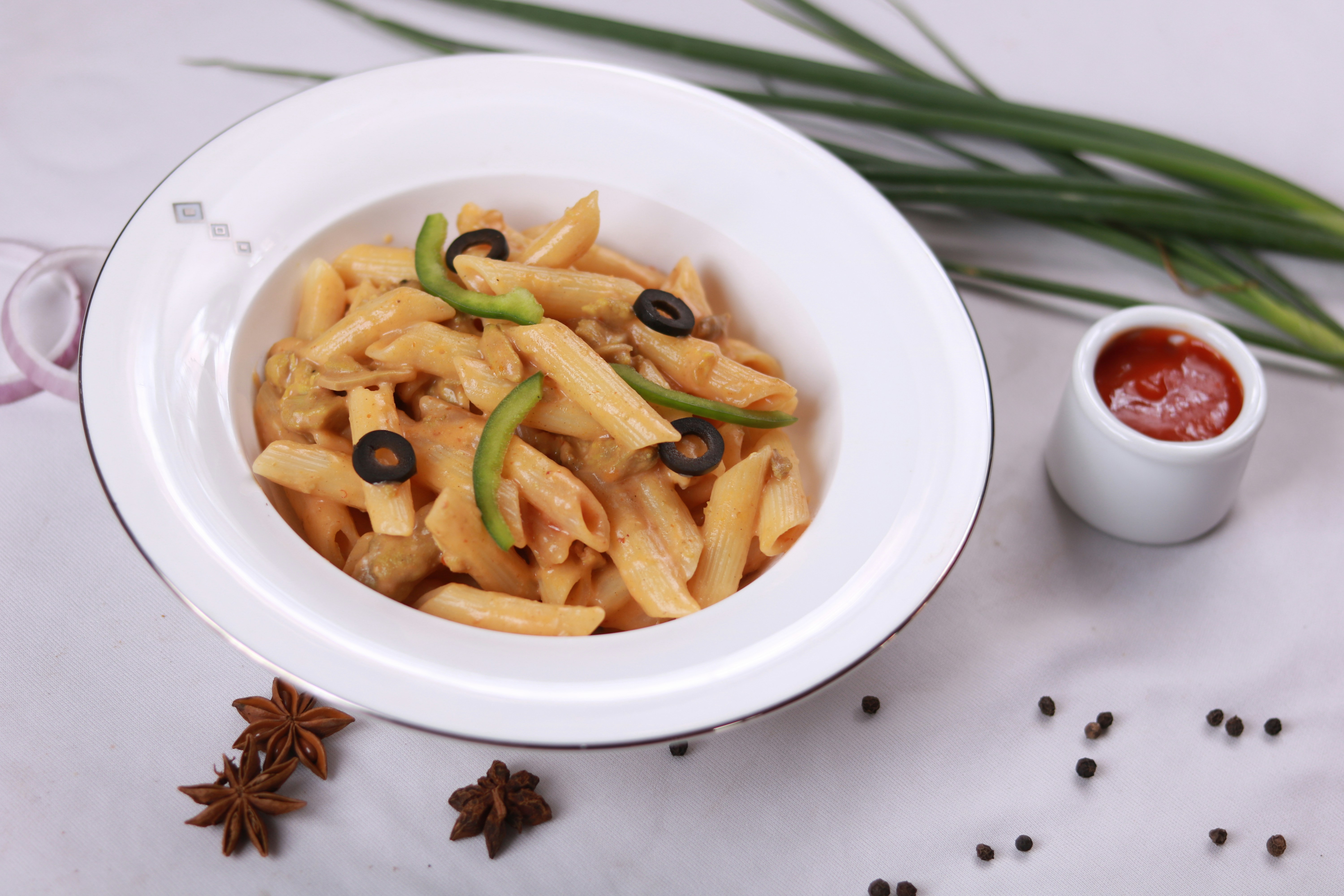 Delicious penne pasta garnished with green peppers and olives, accompanied by a small bowl of sauce. Fresh green onions and spices add a touch of flair.