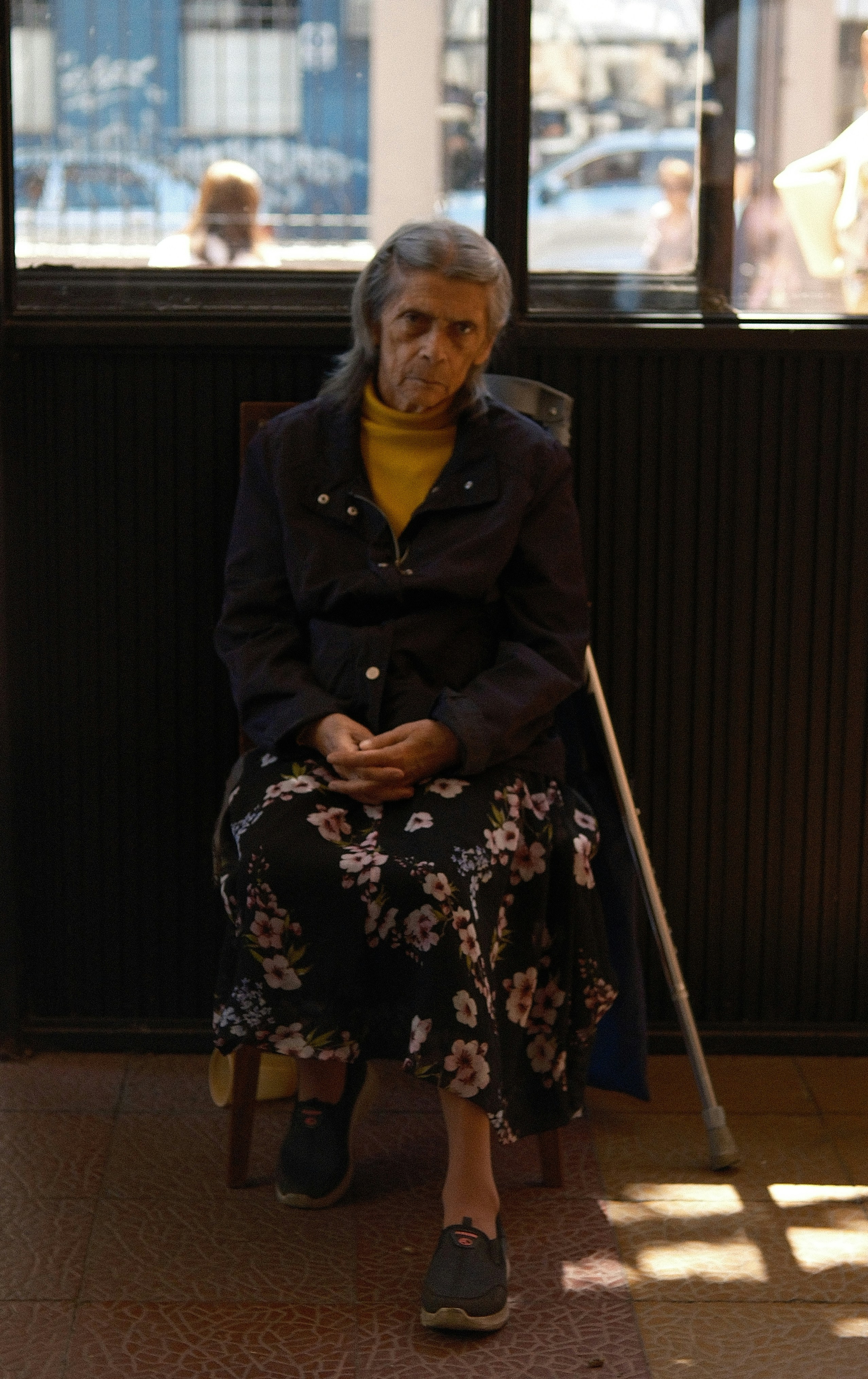 Elderly woman seated in a sunlit room, wearing a dark coat and floral skirt, with a cane resting beside her.