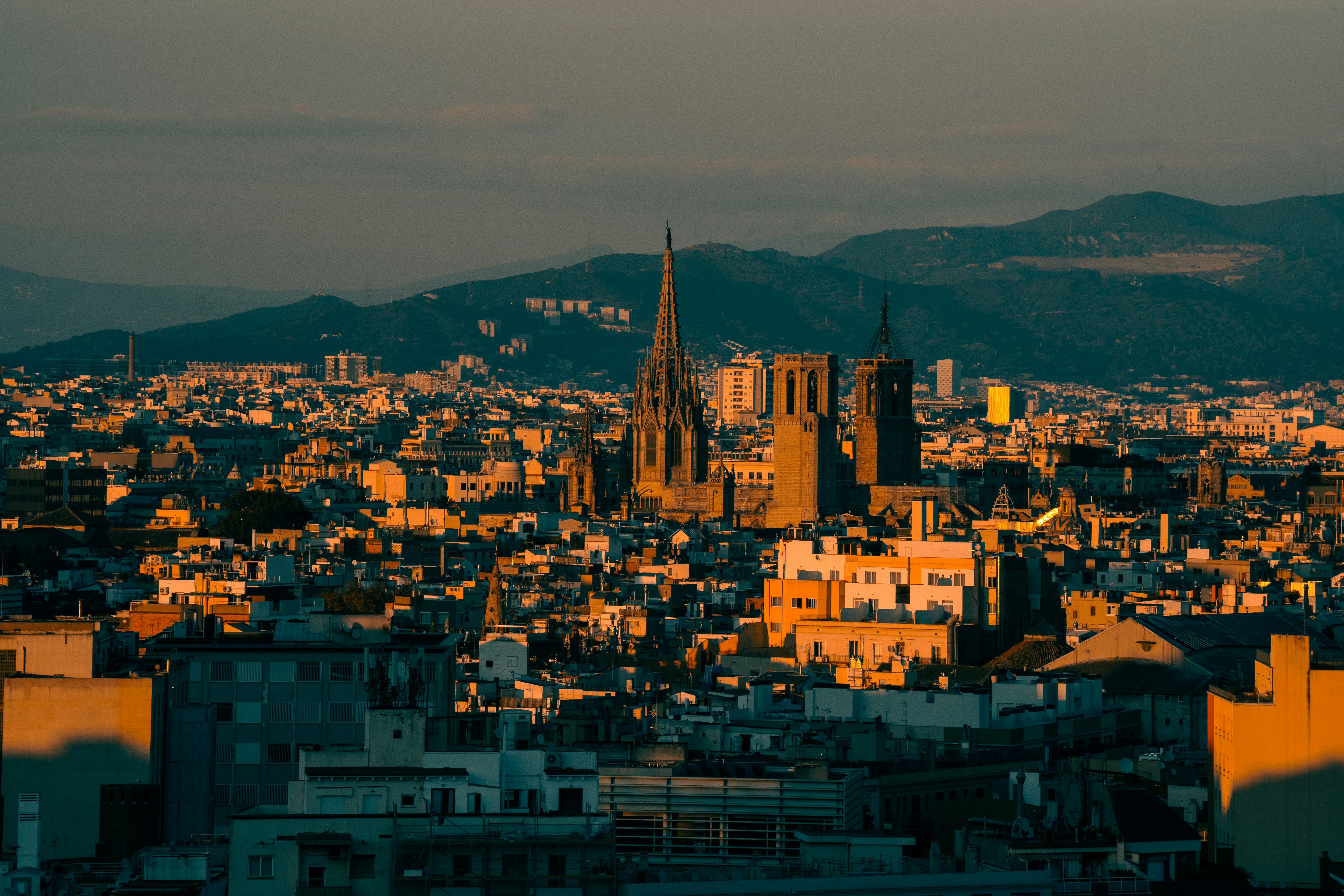 Barcelona cityscape at sunset with prominent cathedral and distant mountains.
