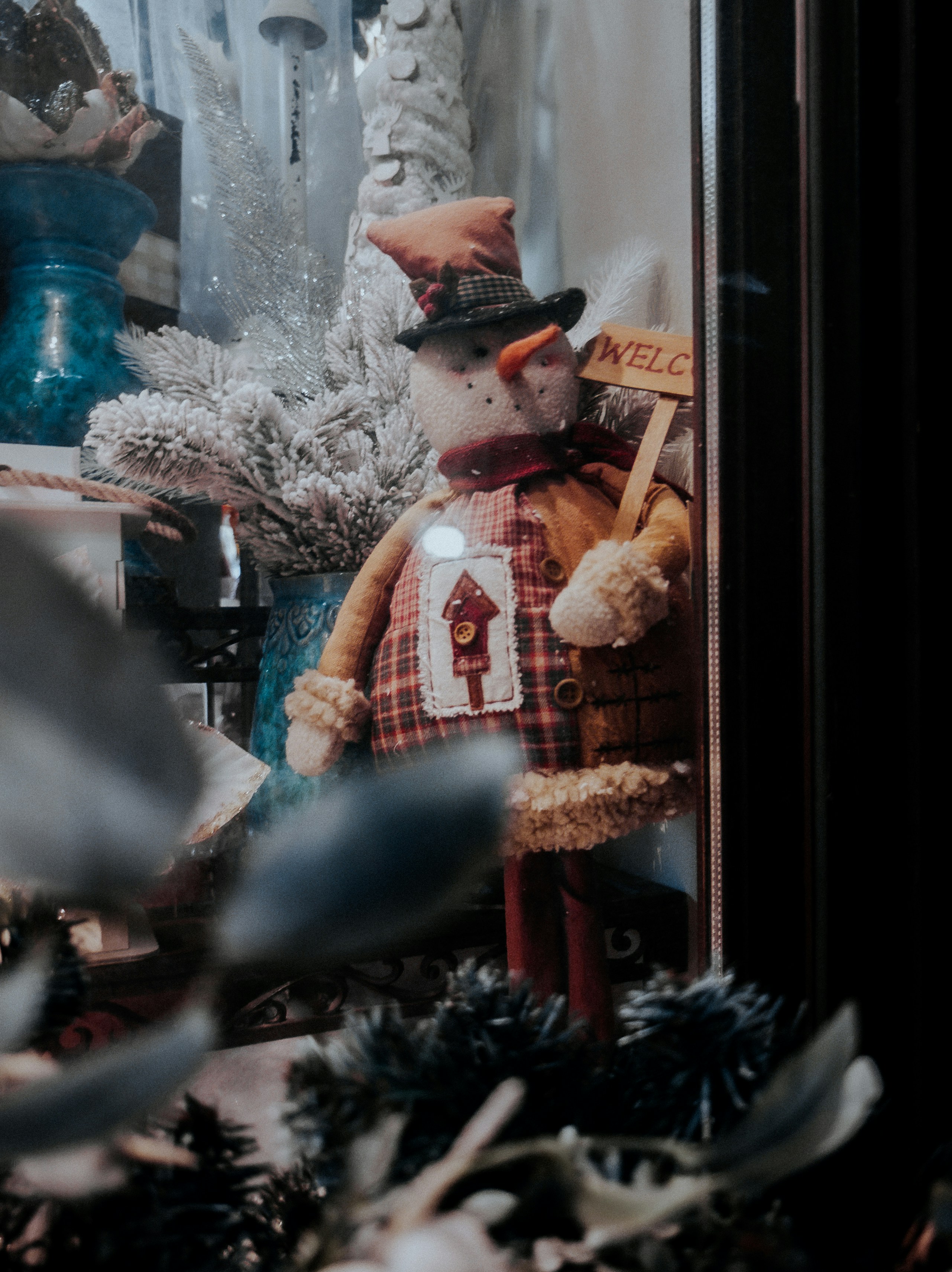 A plush snowman in a plaid outfit stands in a frosted shop window, surrounded by holiday decor and soft lighting.