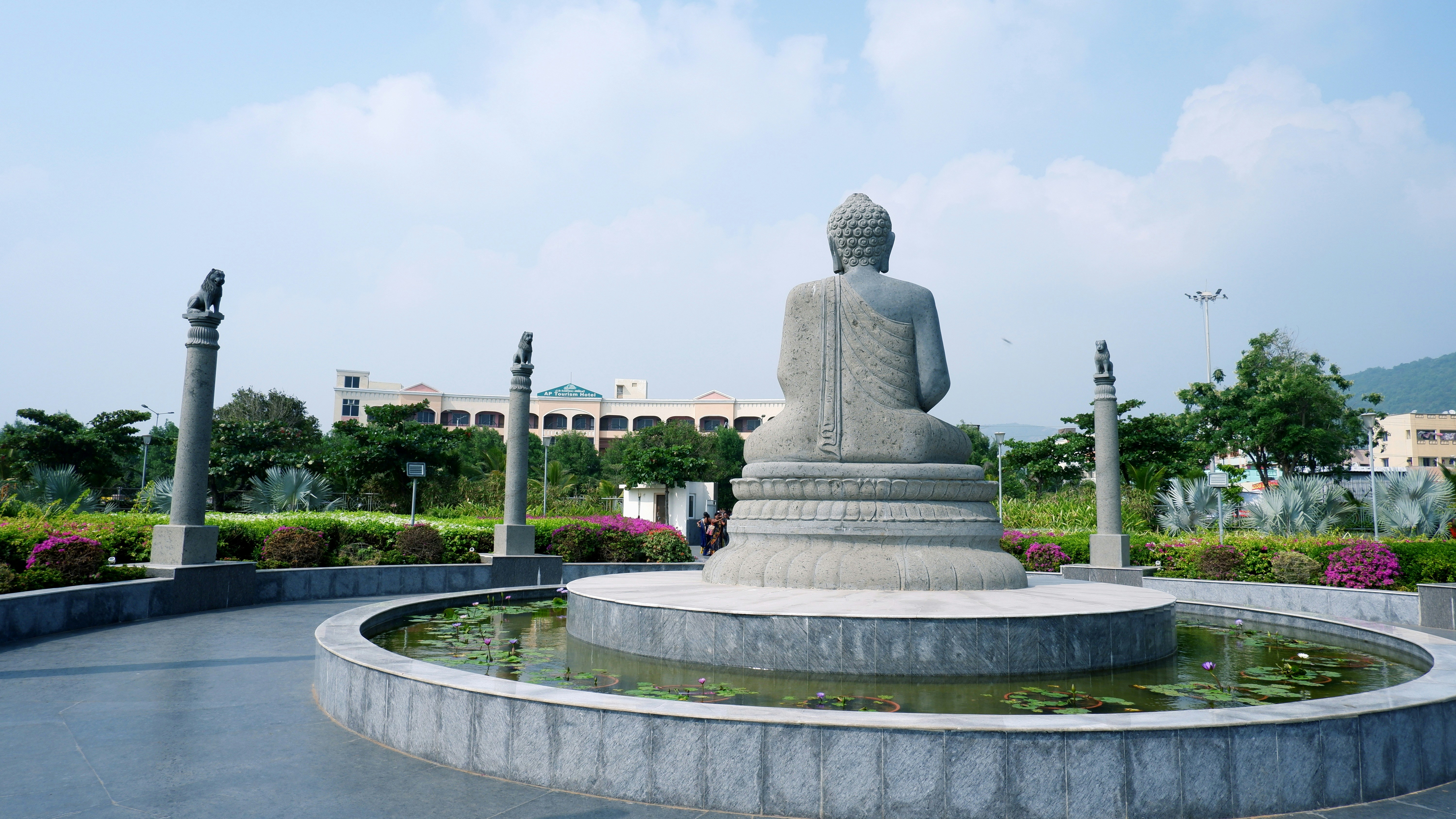 Back view of a large Buddha statue surrounded by a circular pond with lotus flowers, set against a backdrop of lush greenery and distant buildings.