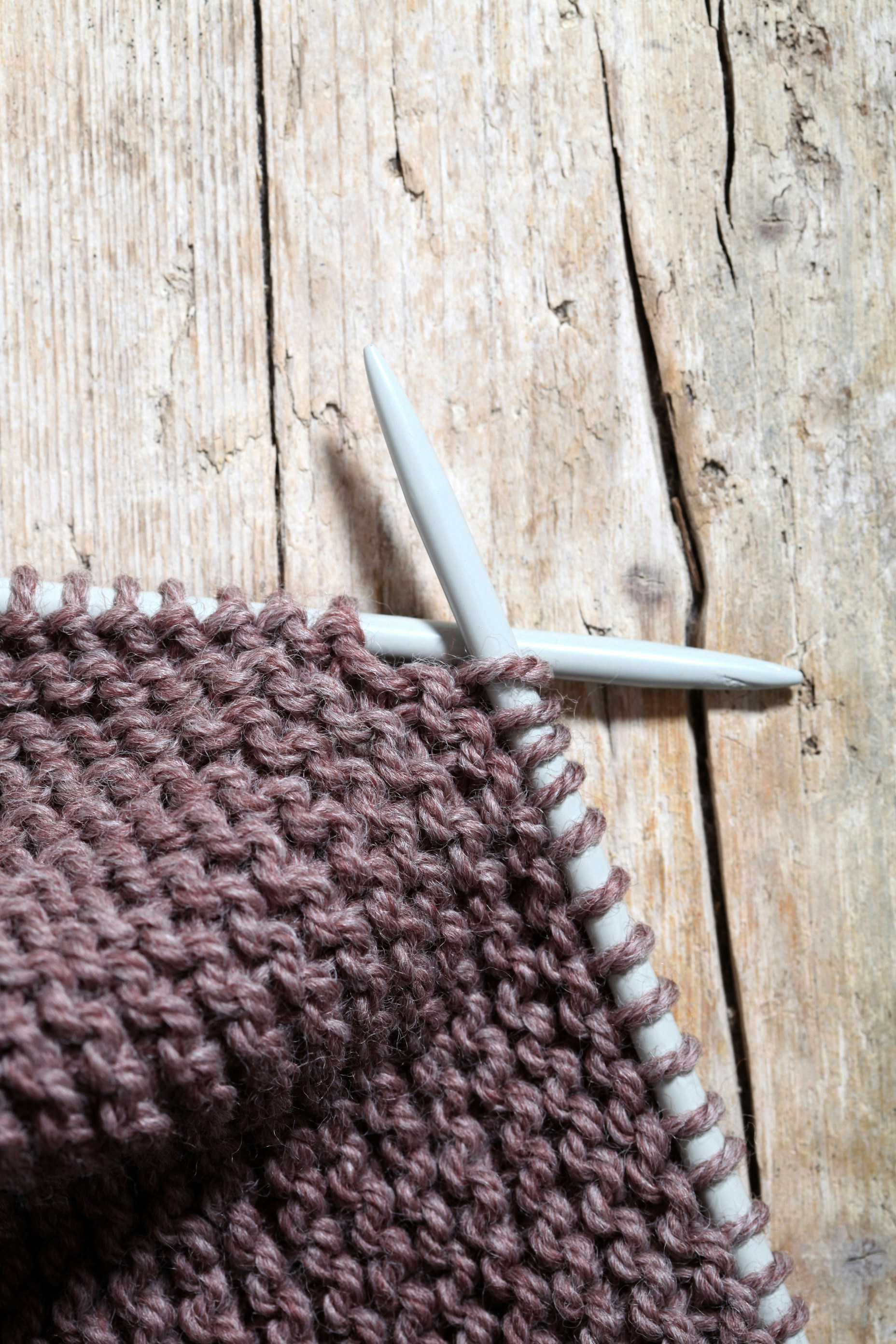 Crossed light-gray needles cradle a mauve knit on weathered wooden planks