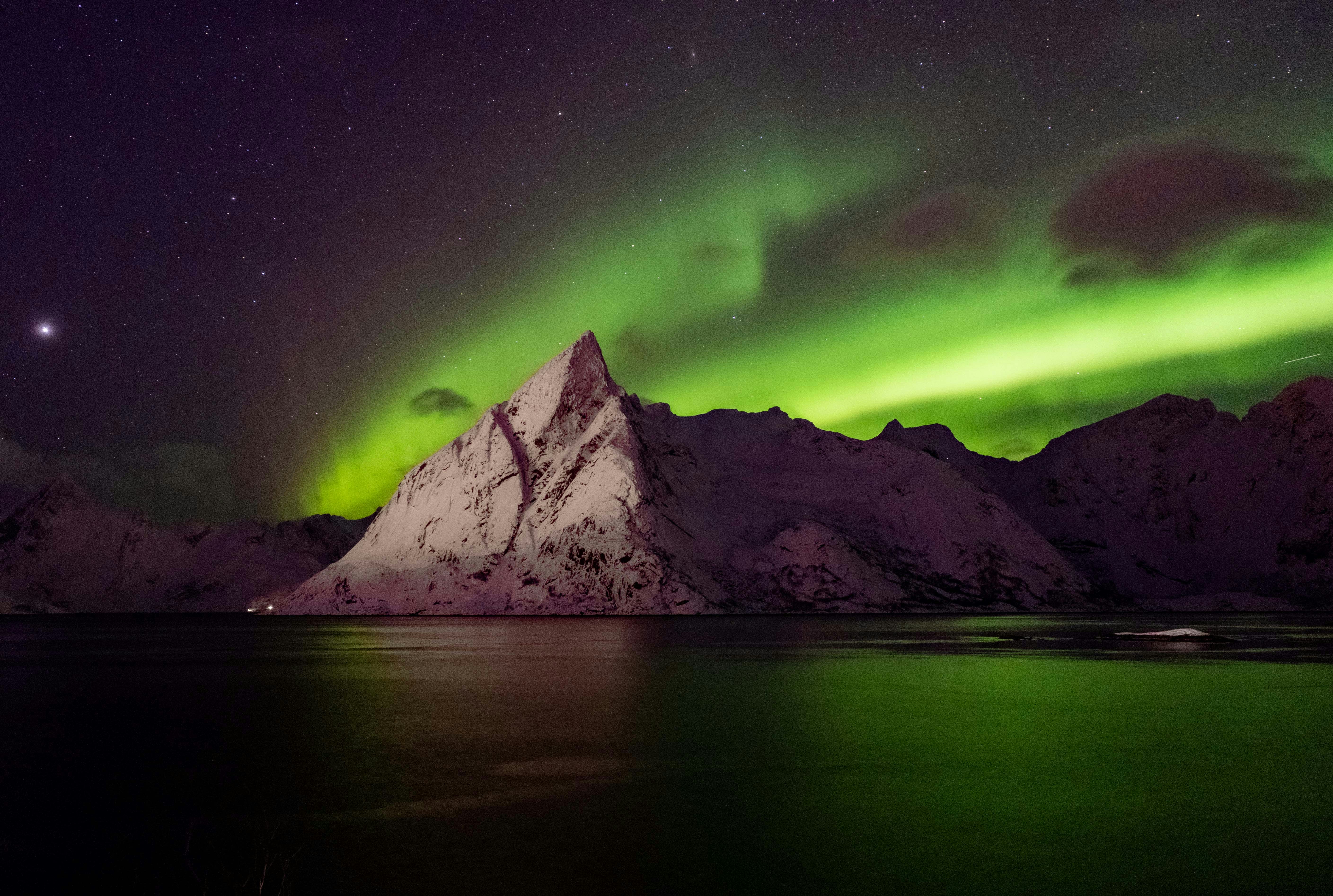 Green aurora dances over snow-covered mountain peaks reflecting in calm water at night.