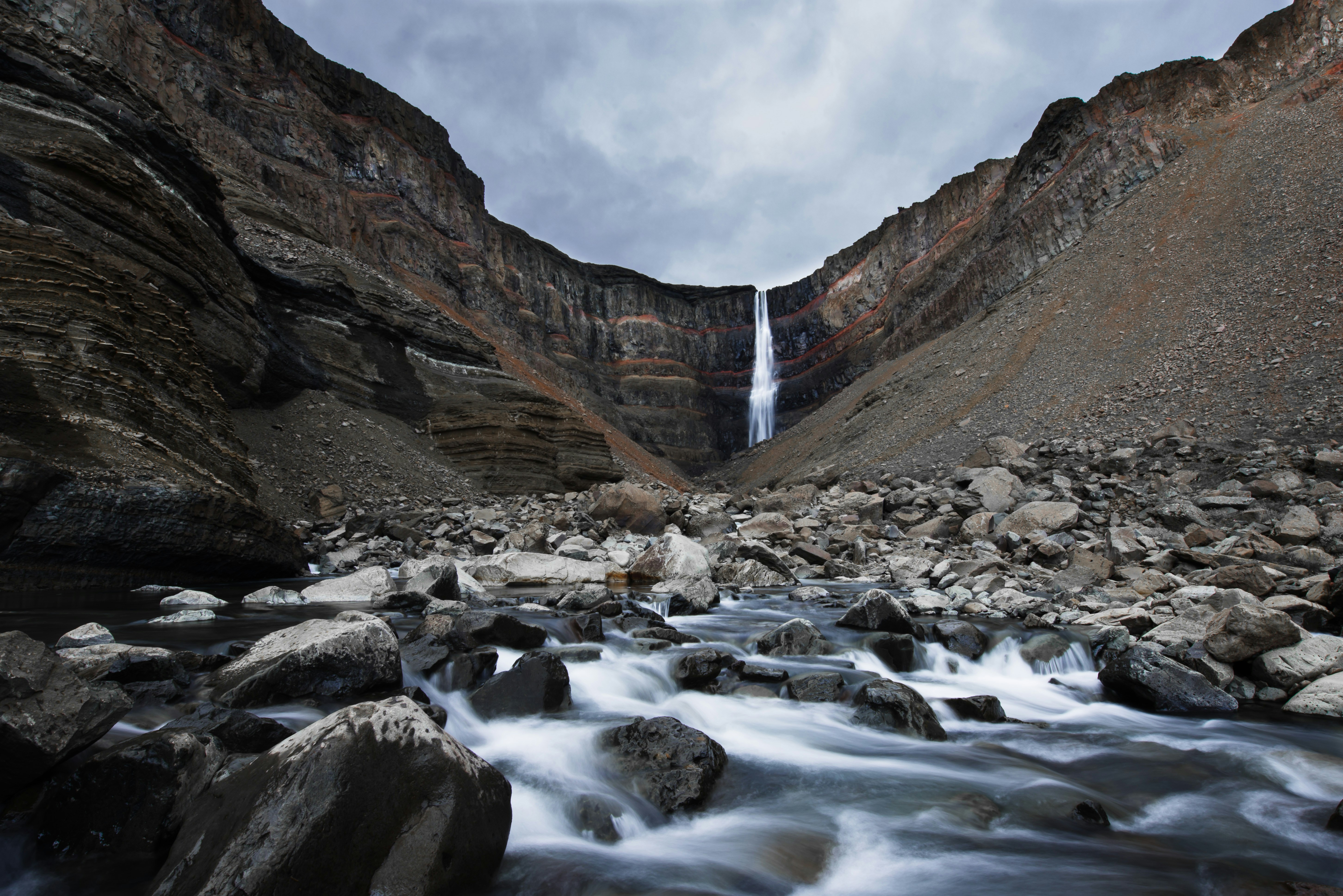 Waterfall tumbling over layered cliffs into a rocky riverbed under a cloudy sky.