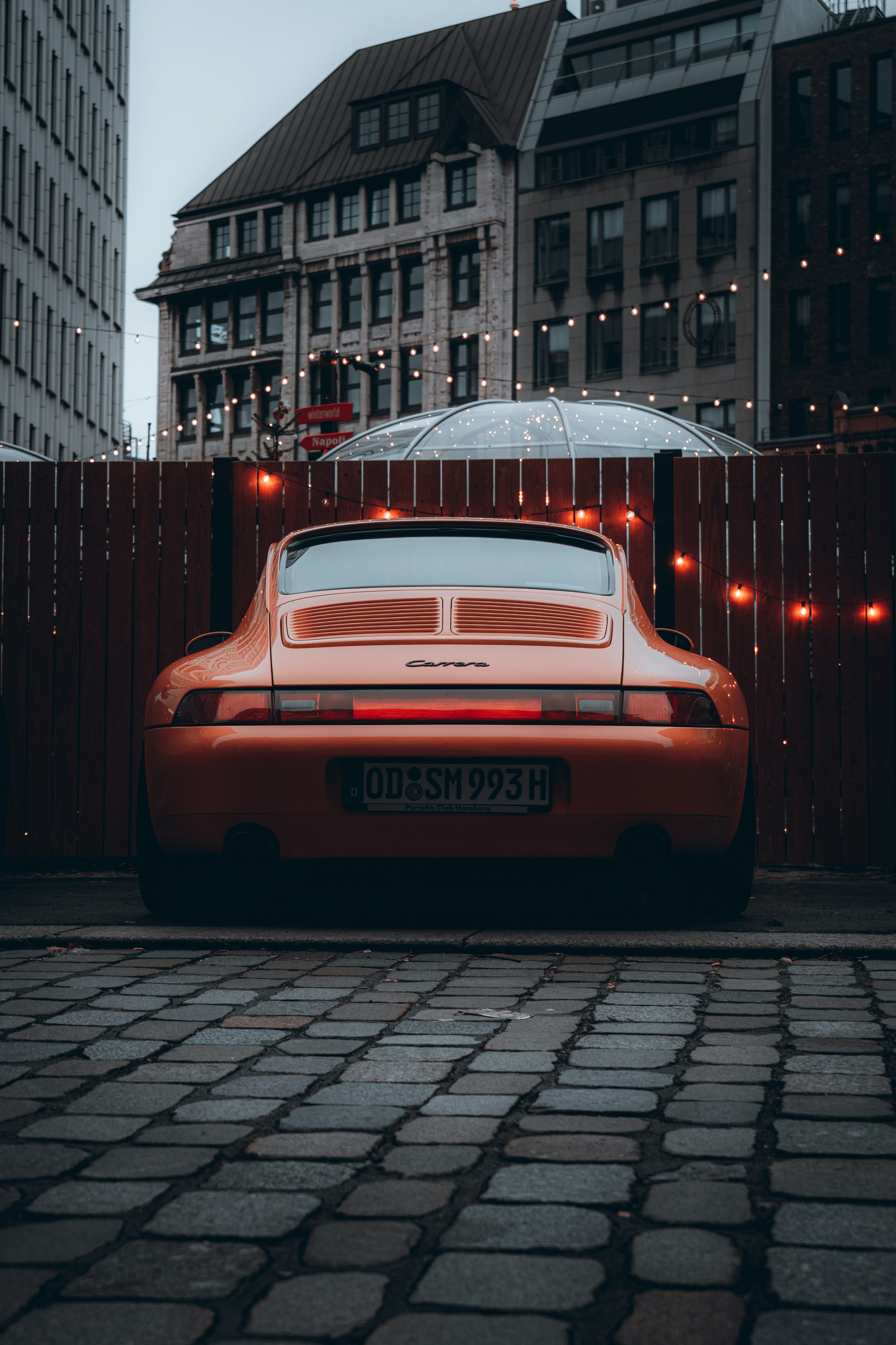 Rear view of an orange Porsche parked behind a string-lit wooden fence along a cobblestone street at dusk.