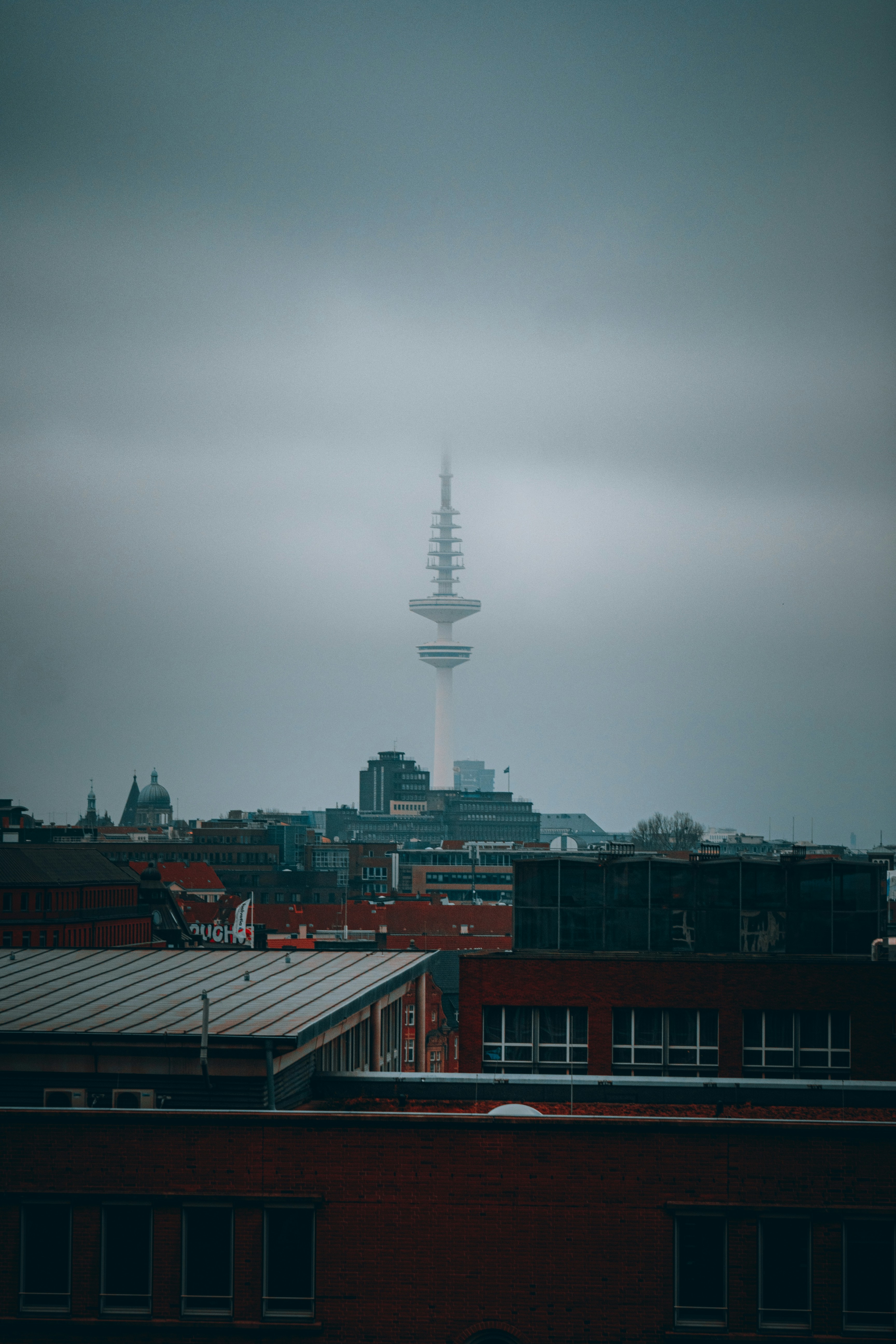 City skyline photograph capturing the Fernsehturm piercing the low clouds above red-brick rooftops.
