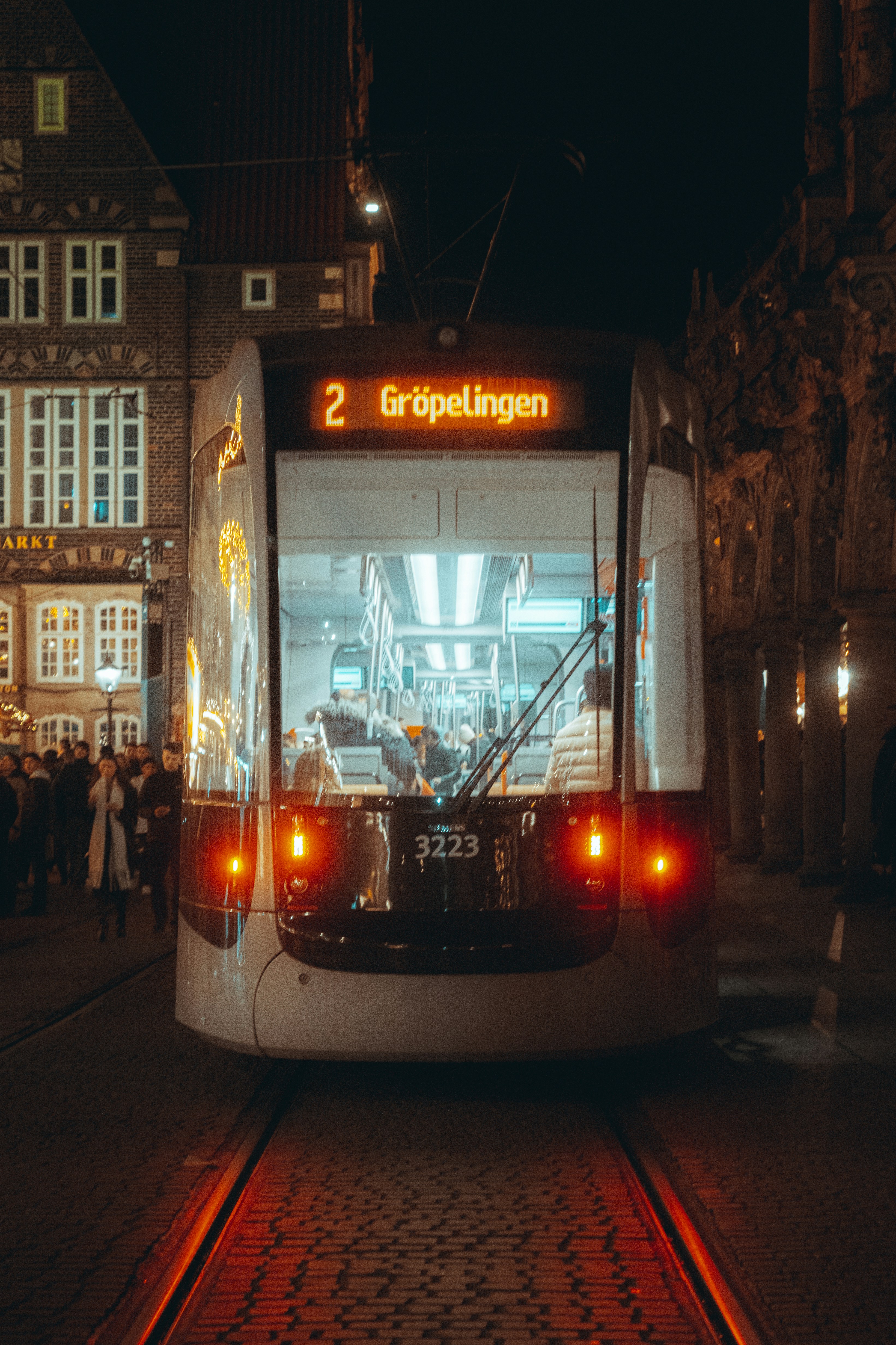 Nocturnal tram with bright interior lighting and red taillights glides along cobblestone rails, passengers visible inside, framed by historic buildings.