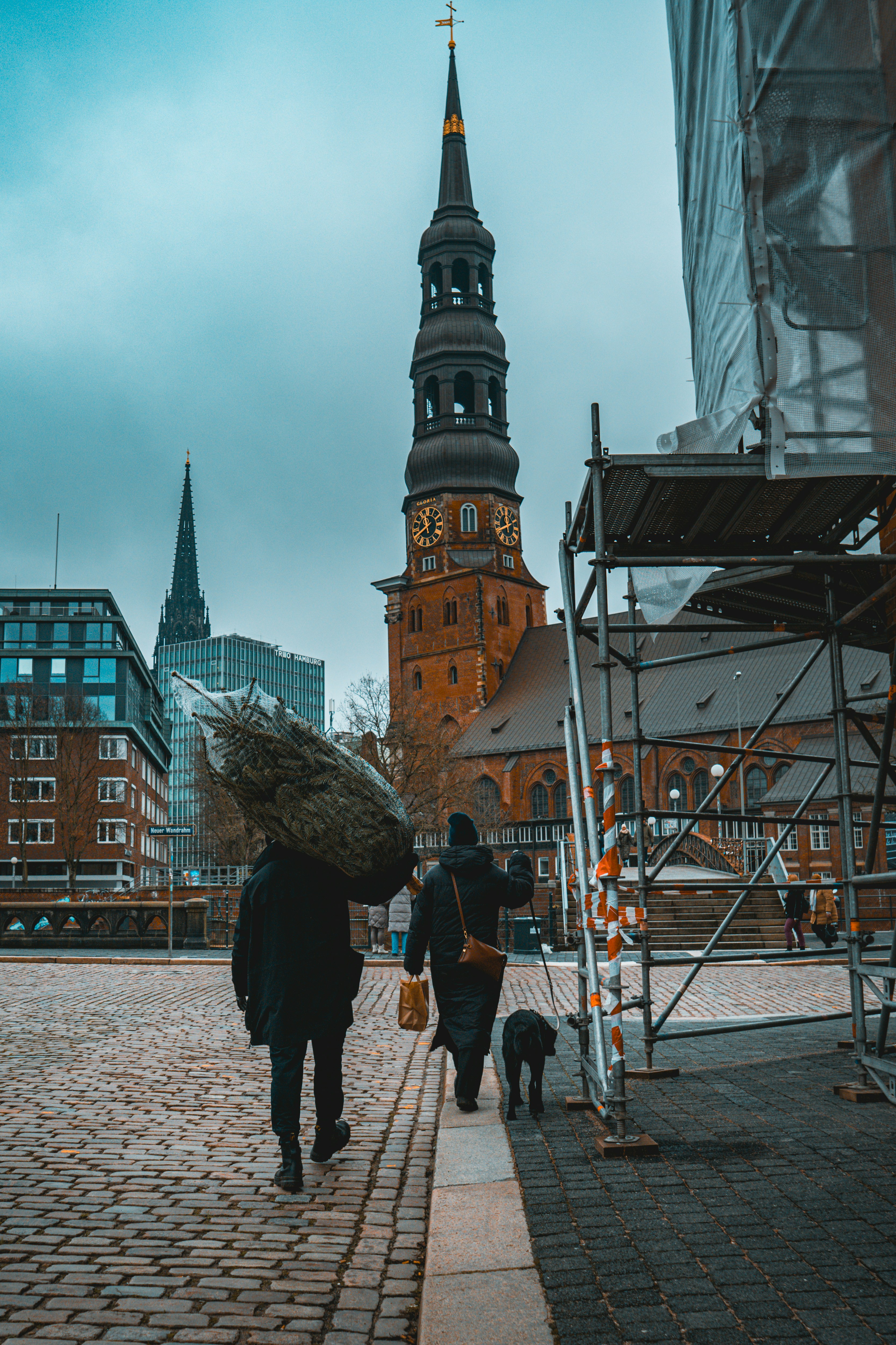 Cobblestone street scene with pedestrians and umbrellas beside a tall brick clock tower, framed by scaffolding under a cloudy sky. Historic architecture meets ongoing construction in a moody urban moment.