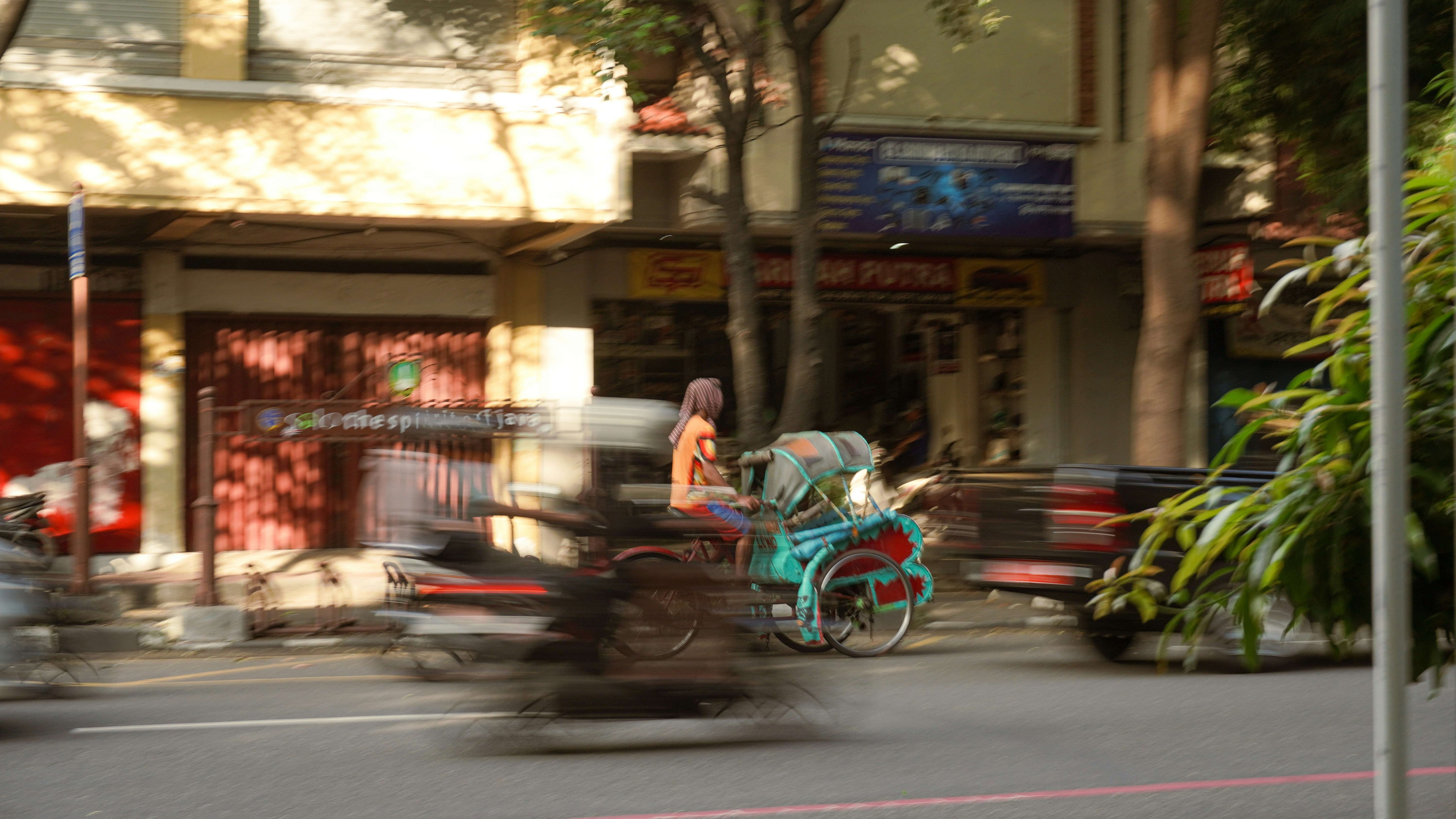 This photo shows a pedicab driver in the middle of a busy city street. His pedicab is brightly colored, with a combination of blue and red, while the driver is wearing casual clothes with a head covering. Motorized vehicles speed around him, creating a blurry effect that shows the dynamics and busyness of the city. Amid the hustle and bustle of the city filled with fast-moving vehicles, a pedicab driver continues to drive slowly, as if he is a symbol of calm amidst the rapid flow of modernity. | A blurry photo of a person riding a bike down a street