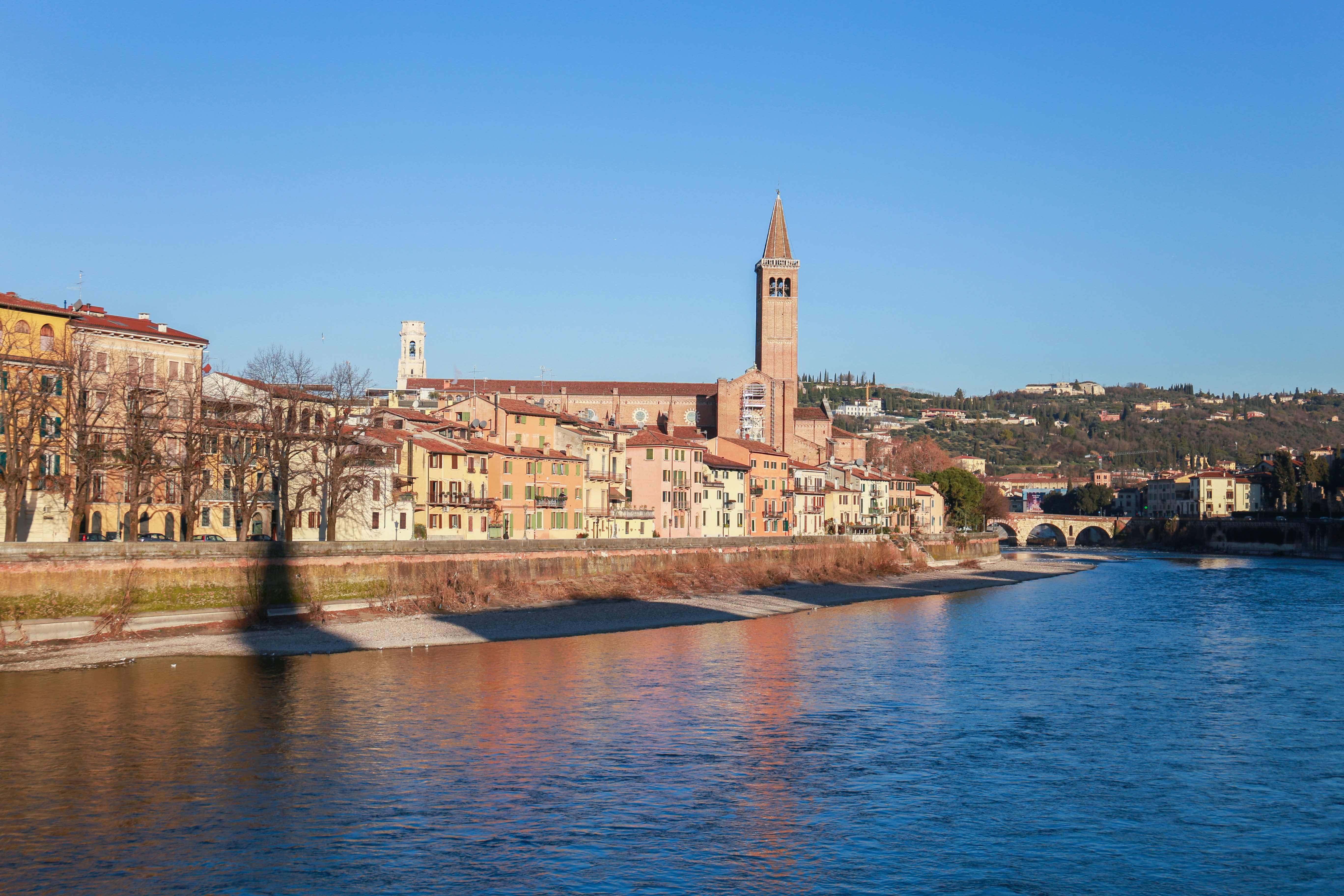 Sunlit riverside townscape with historic buildings and a prominent tower under a clear blue sky.