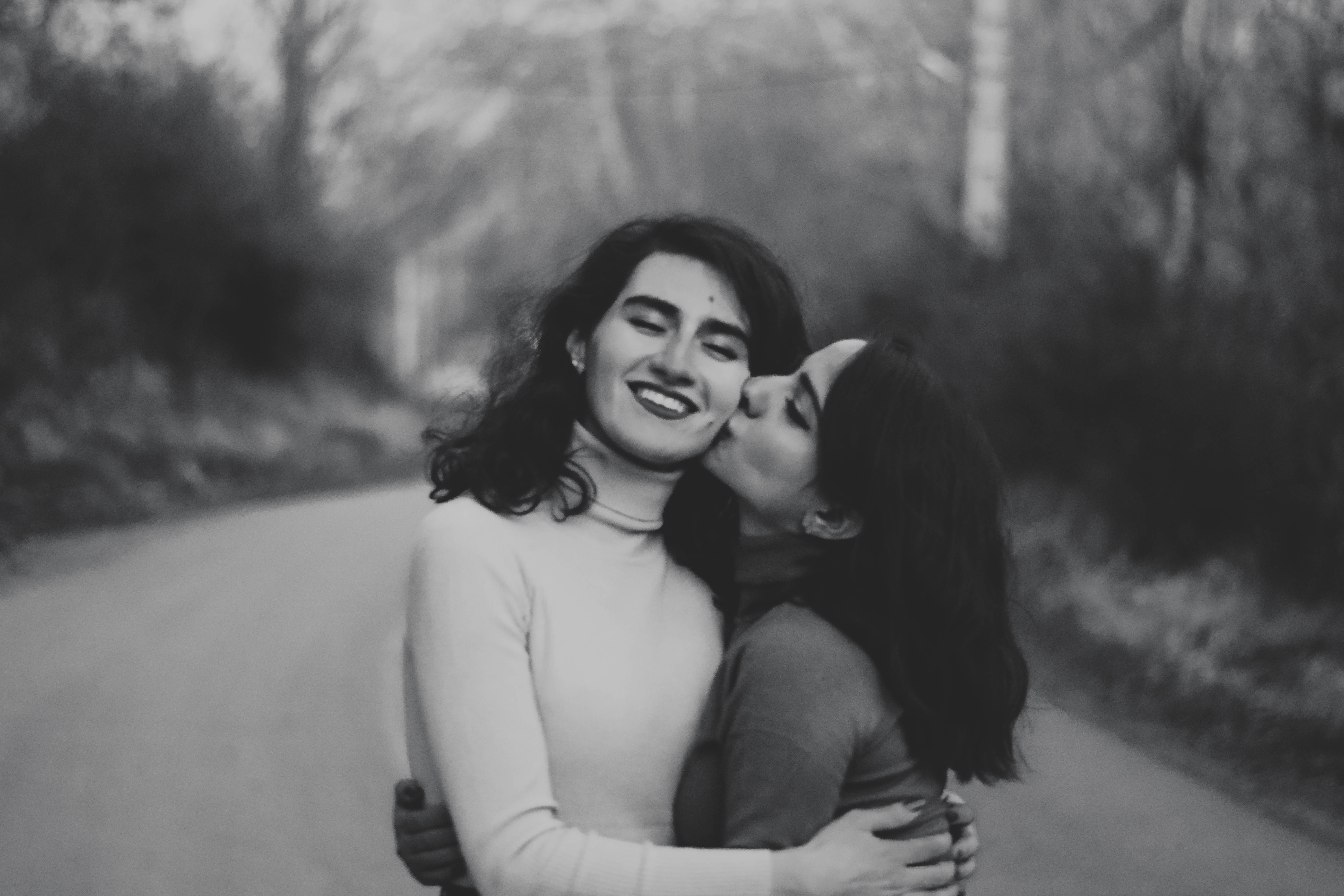 Two women embracing on a deserted road in black and white, conveying warmth and connection in a blurred natural setting.