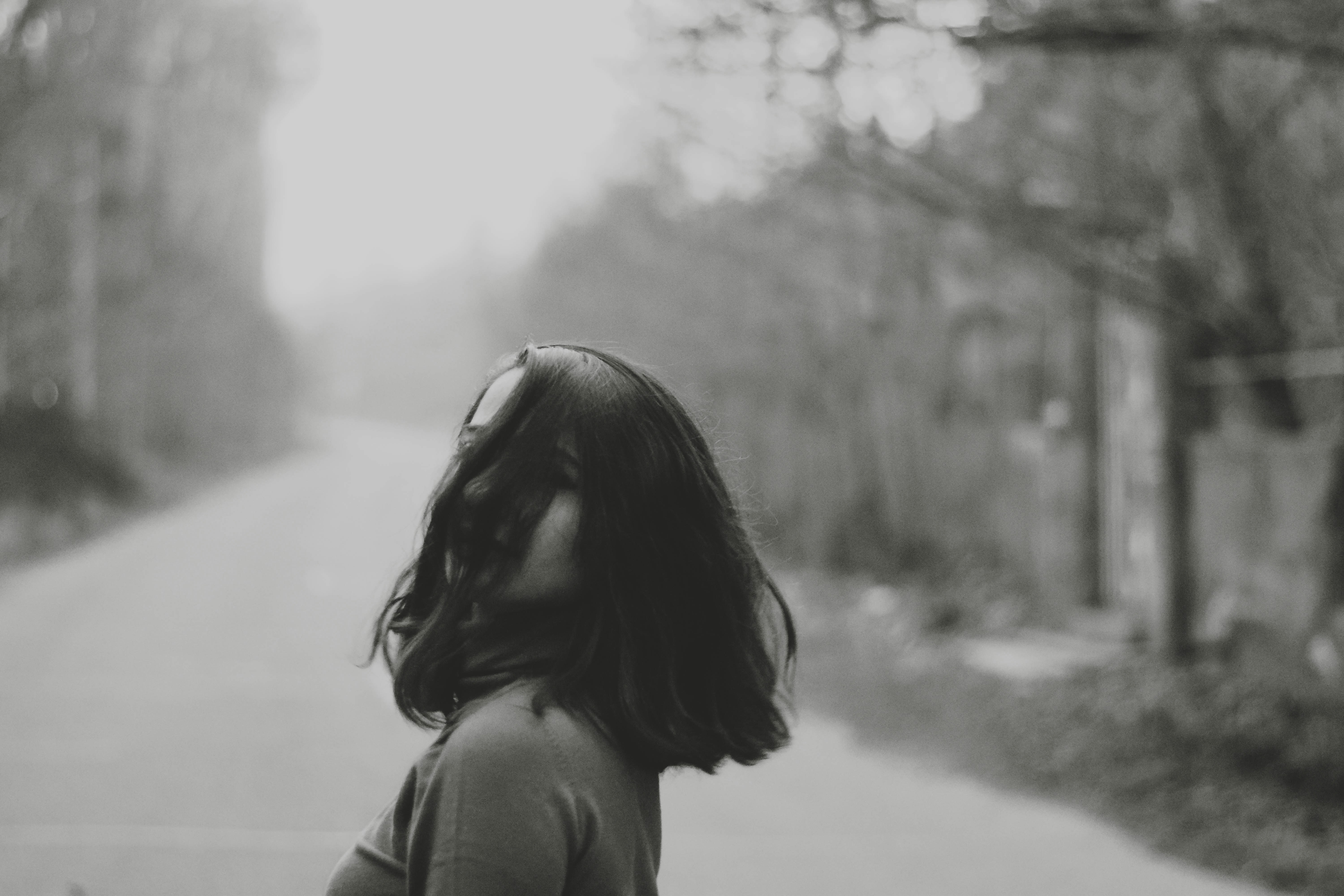 Woman walks along a misty road, her hair caught in motion against blurred trees and an empty street. Monochrome tones enhance the tranquil atmosphere.