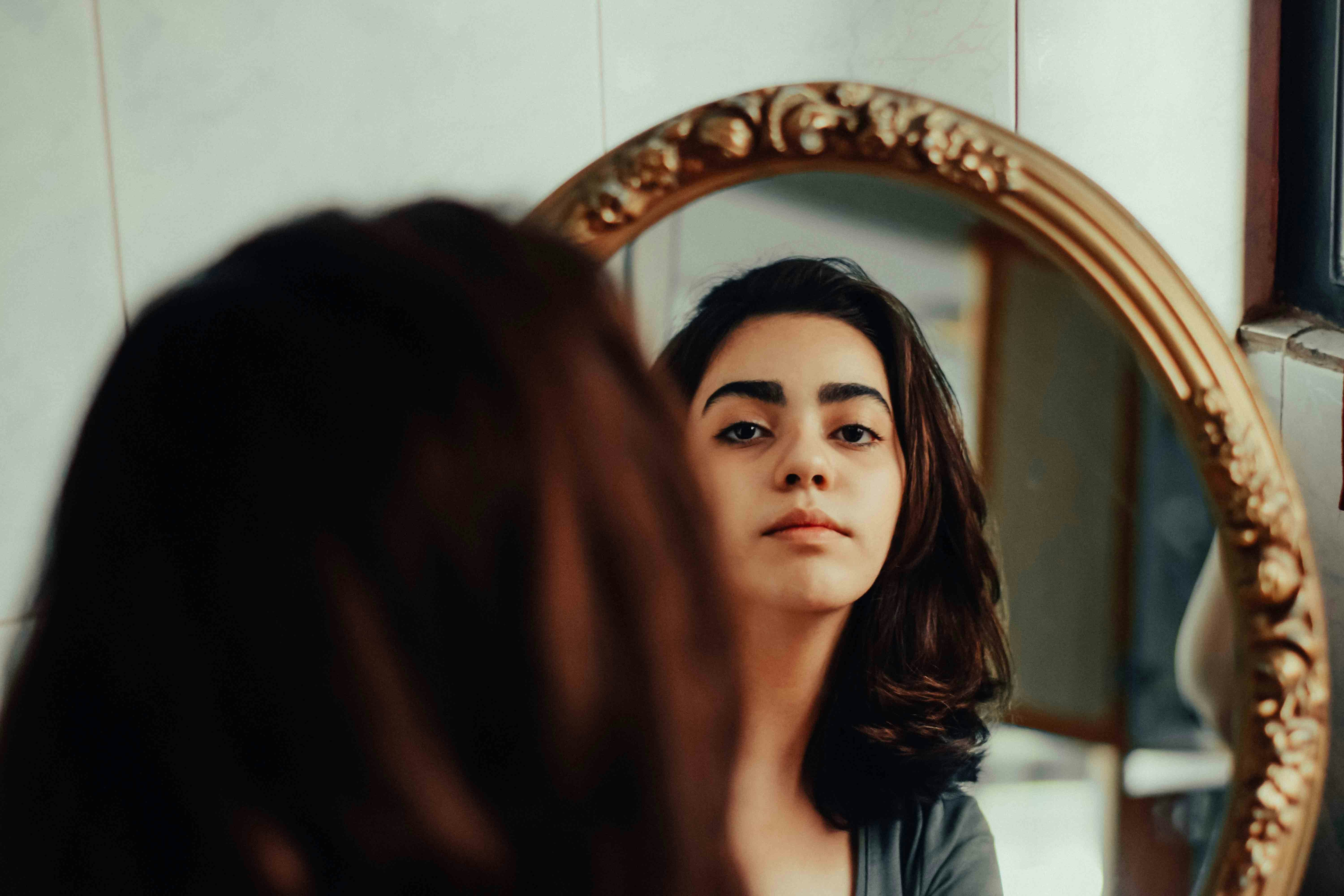 Woman gazes at her reflection in a gold-framed mirror, surrounded by soft lighting and gentle shadows.
